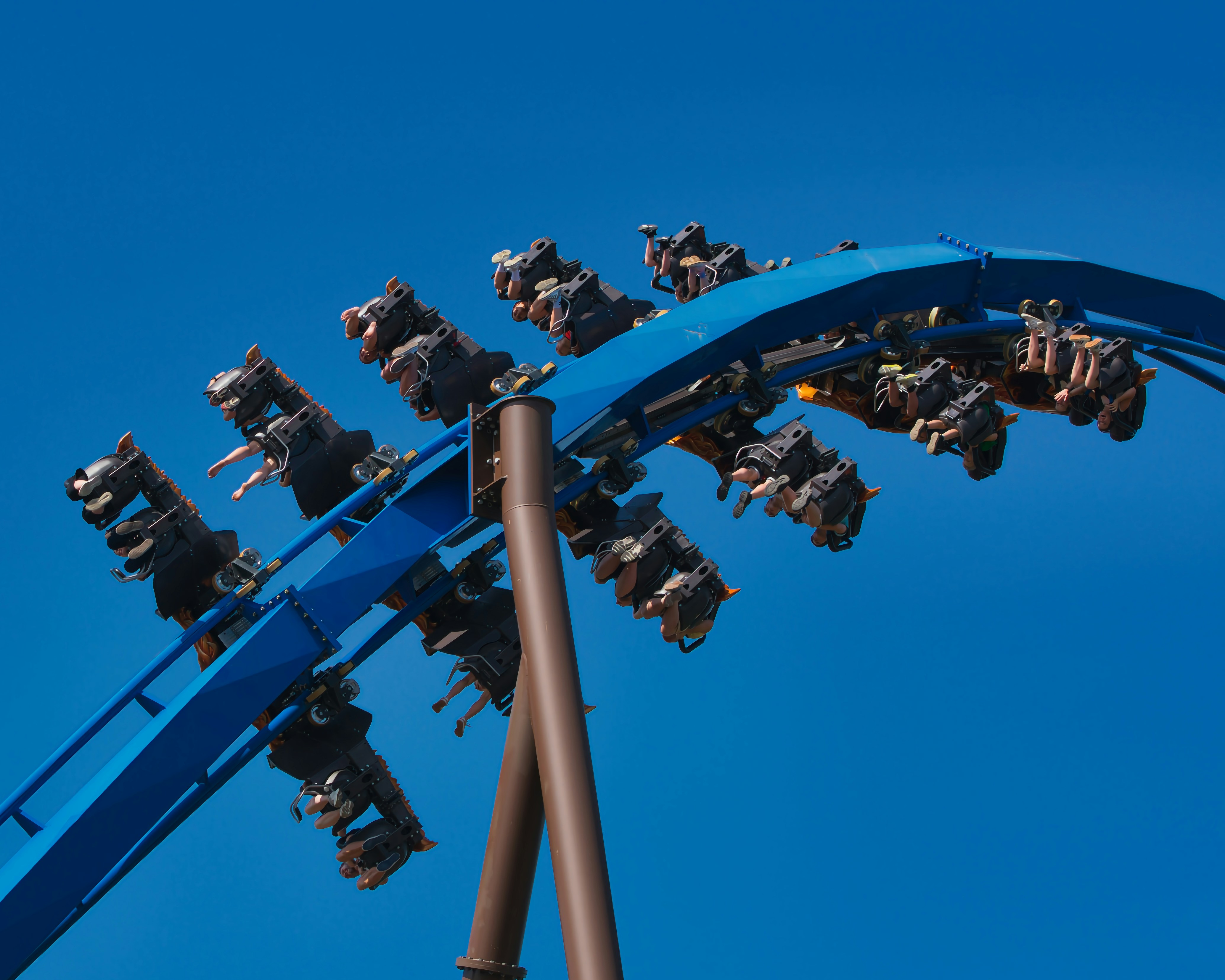 Fēnix — the phoenix takes flight. From a 40 m lift hill into a dive drop, this B&M Wing Coaster whips you through three inversions (Immelmann, Zero‑G roll, dive drop) at 95 km/h under fiery wings and enchanting Avalon theming. Type: Wing Coaster (Bolliger & Mabillard) Opened: 7 July 2018 Height: 40 m Length: 813 m Top Speed: 95 km/h Inversions: 3 Duration: ~1:45 min G‑Force: 5 g Trains: 2 trains with 6 cars (4 riders per car) | People are riding a roller coaster on a blue track.