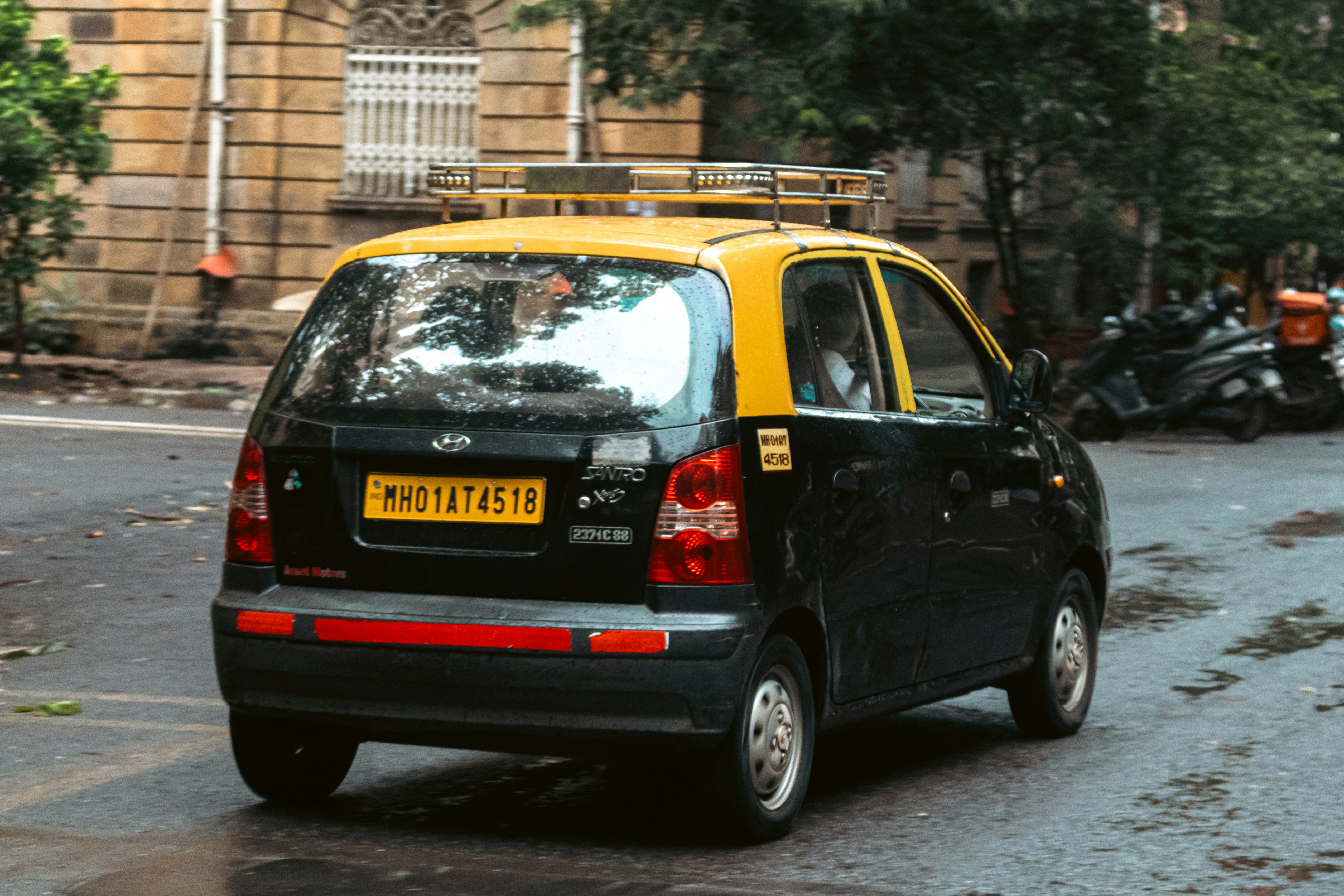 A black and yellow Hyundai Santro taxi drives through the streets of Ballard Estate, Mumbai, on a Sunday July evening. | A yellow-top taxi waits on a city street.