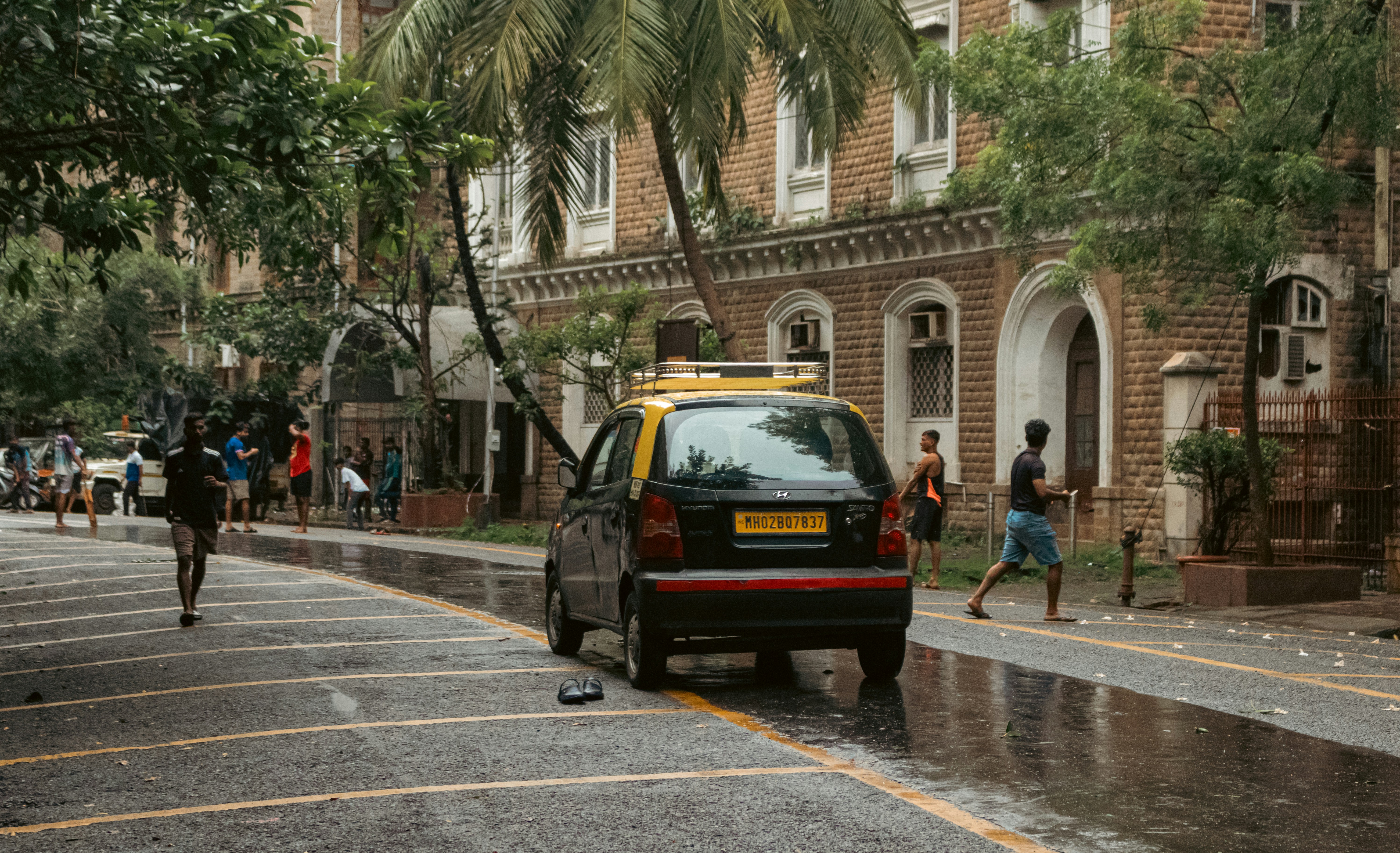 A black taxi parked on a rain-soaked street, surrounded by lush greenery and people enjoying the weather. The scene captures a vibrant urban atmosphere.