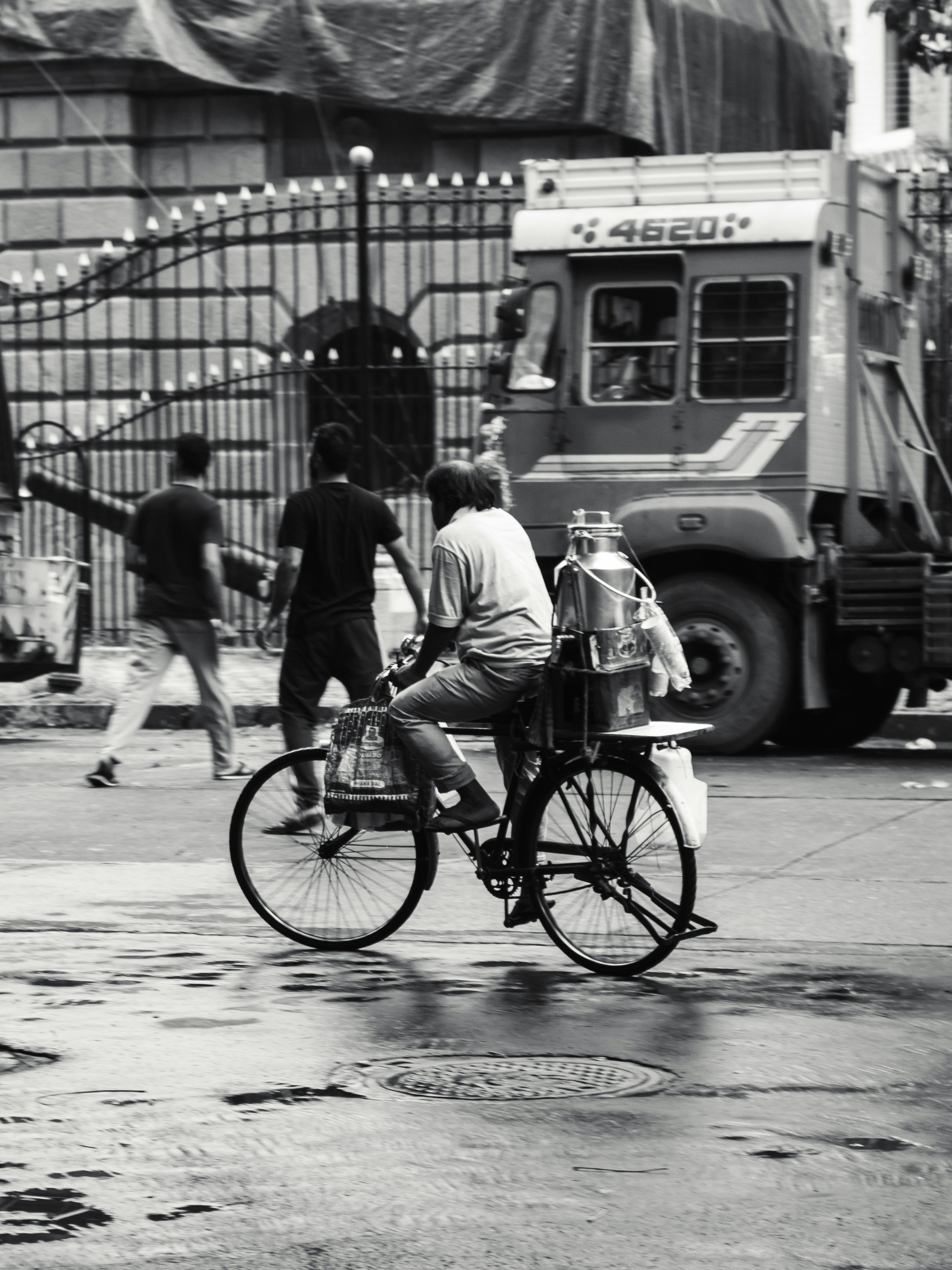 A tea seller cycles down the road with a large thermos strapped to the backseat of his bicycle, embodying a familiar scene from urban India. The image captures the quiet determination of local street vendors who serve countless cups of chai daily. | A man rides a bike with cargo on the street.
