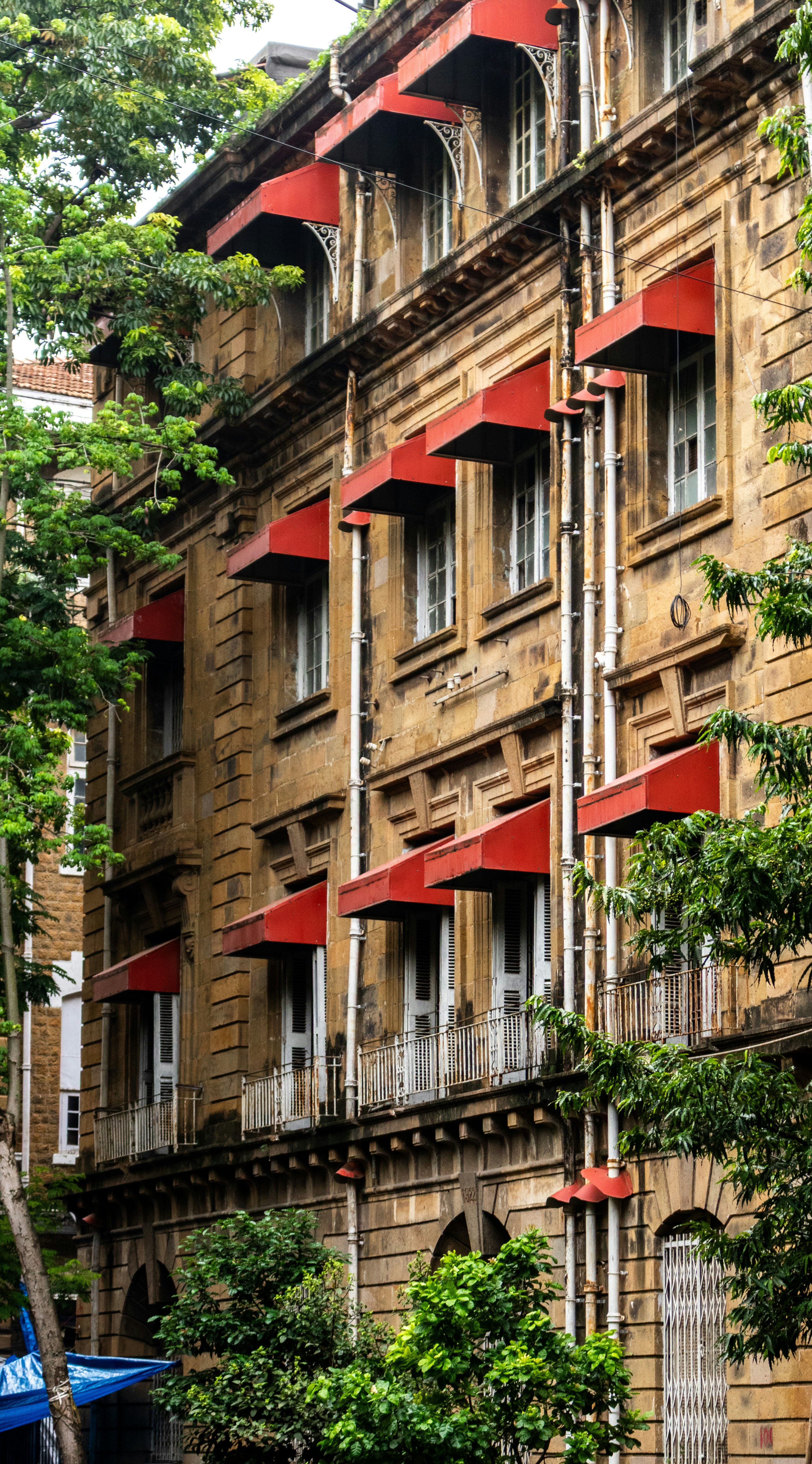 The facade of a heritage building in Ballard Estate, Mumbai. The structure showcases classic colonial design elements such as arched windows, stone masonry, and symmetrical lines. Set in the heart of South Mumbai’s commercial area, the building reflects the city's layered architectural history and urban formality. | Old building with red awnings and leafy surroundings.
