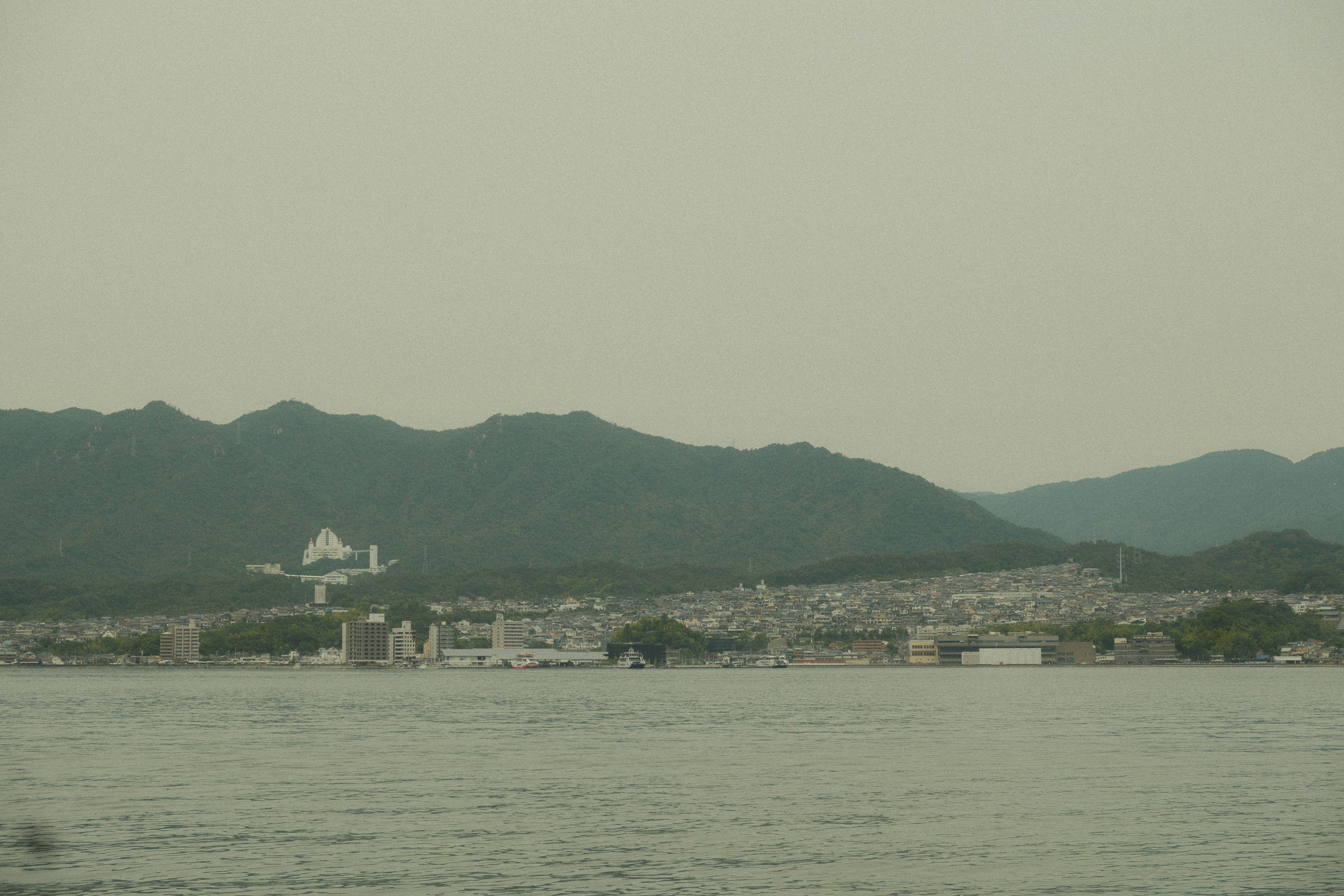A tranquil coastal view showcasing a distant city nestled against lush mountains under a hazy sky.