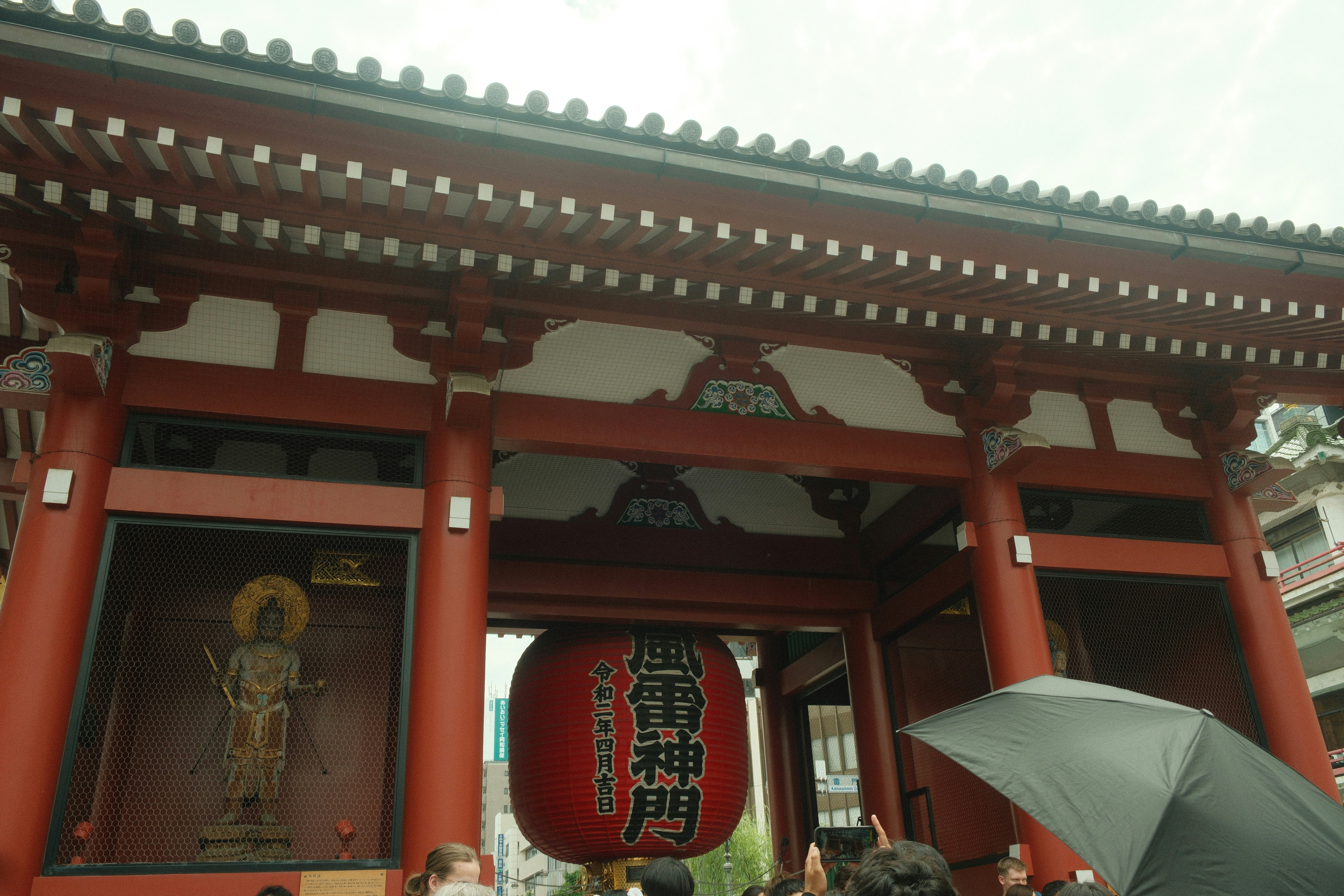 Intricate red wooden structure adorned with traditional motifs, featuring a prominent lantern and a guardian statue at the entrance to a historic site.