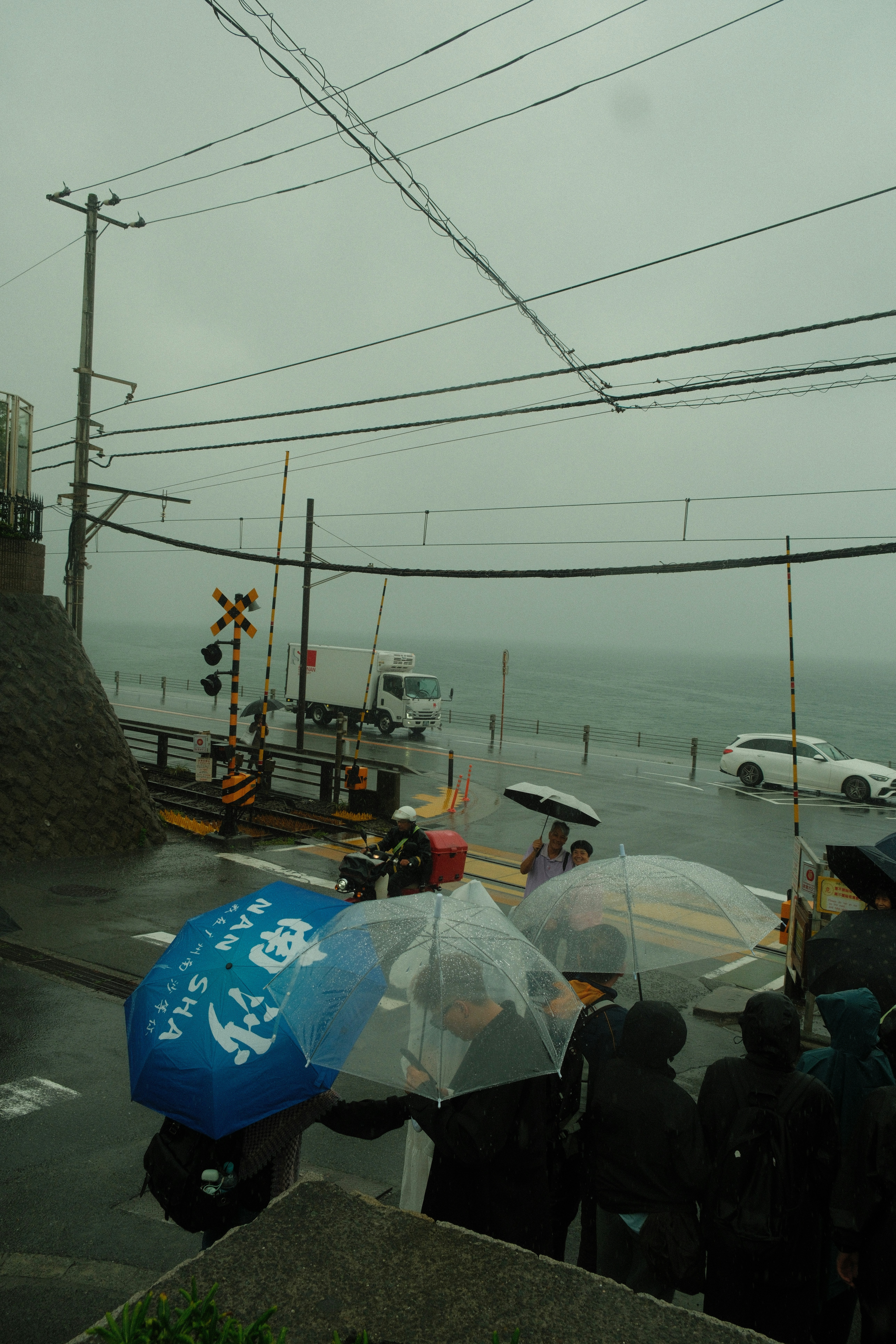 Group of people with umbrellas waiting at a train crossing on a rainy day, with a view of the ocean in the background.