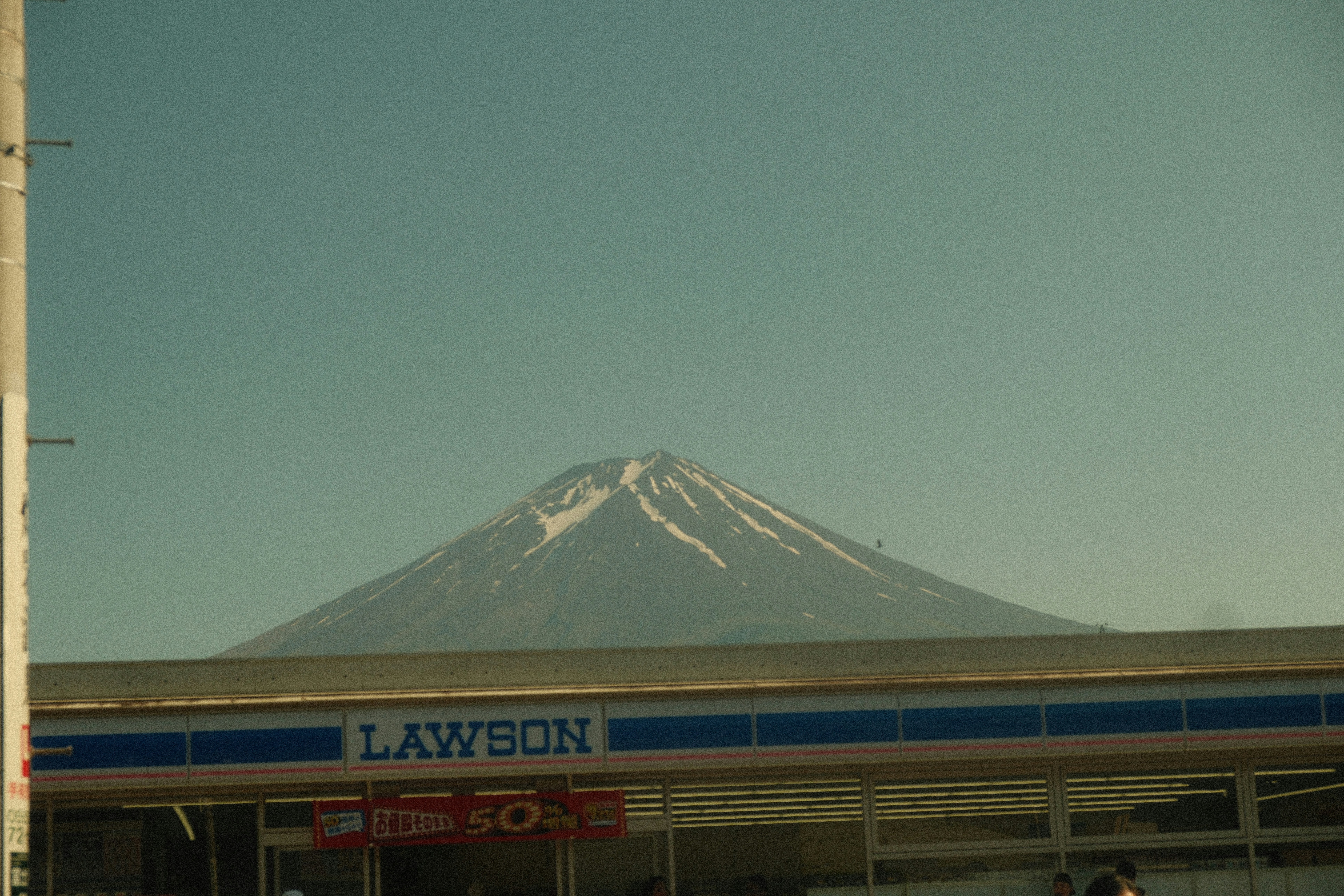 Mount Fuji looms majestically in the background, partially overshadowed by a Lawson convenience store in the foreground. The scene captures a blend of urban life and natural beauty.