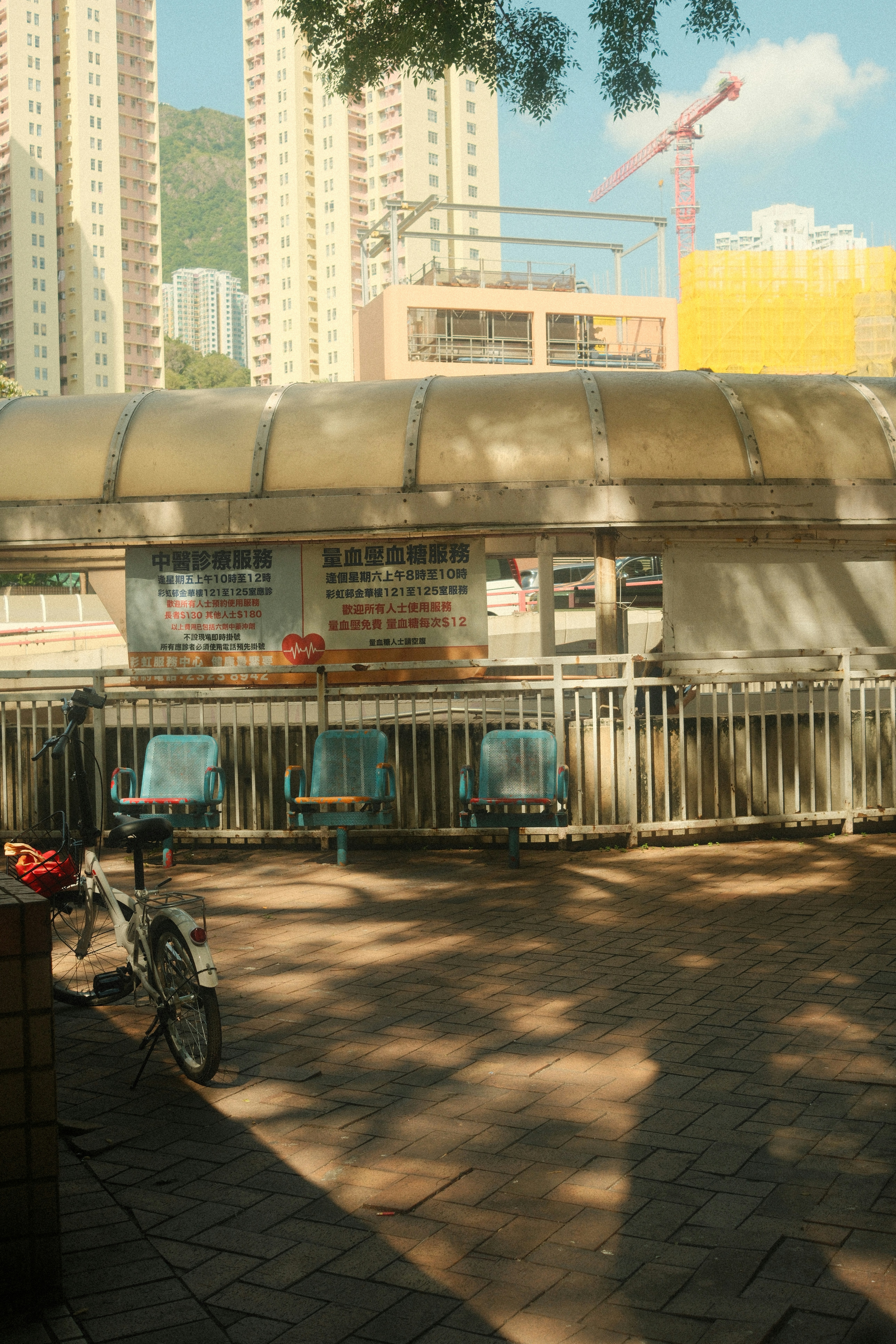 A quiet bus stop with empty blue seats, shadowed by a large overhang, set against a backdrop of towering residential buildings and construction activity.