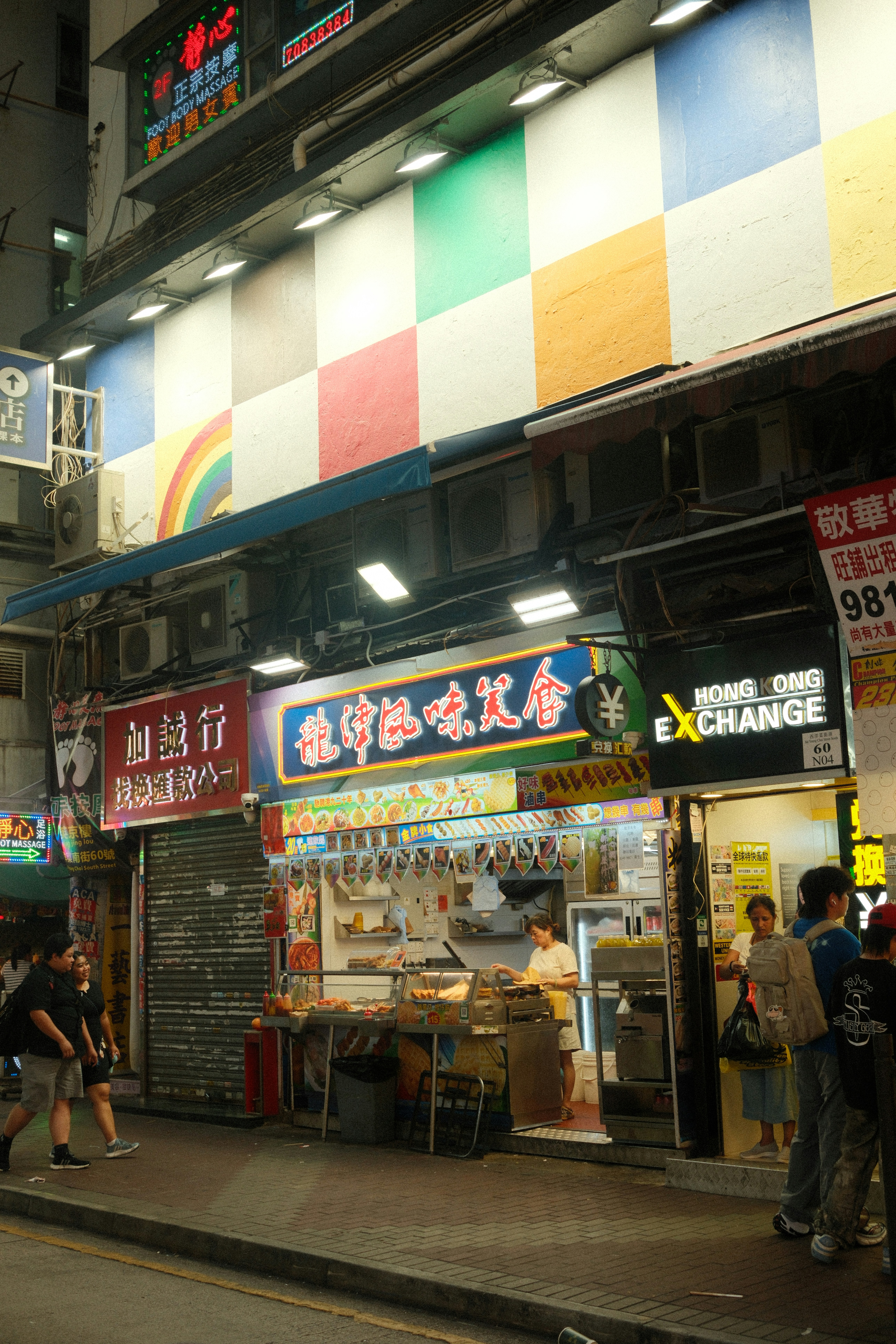 Vibrant street scene featuring a bustling food stall illuminated at night, with colorful signage and pedestrians passing by.