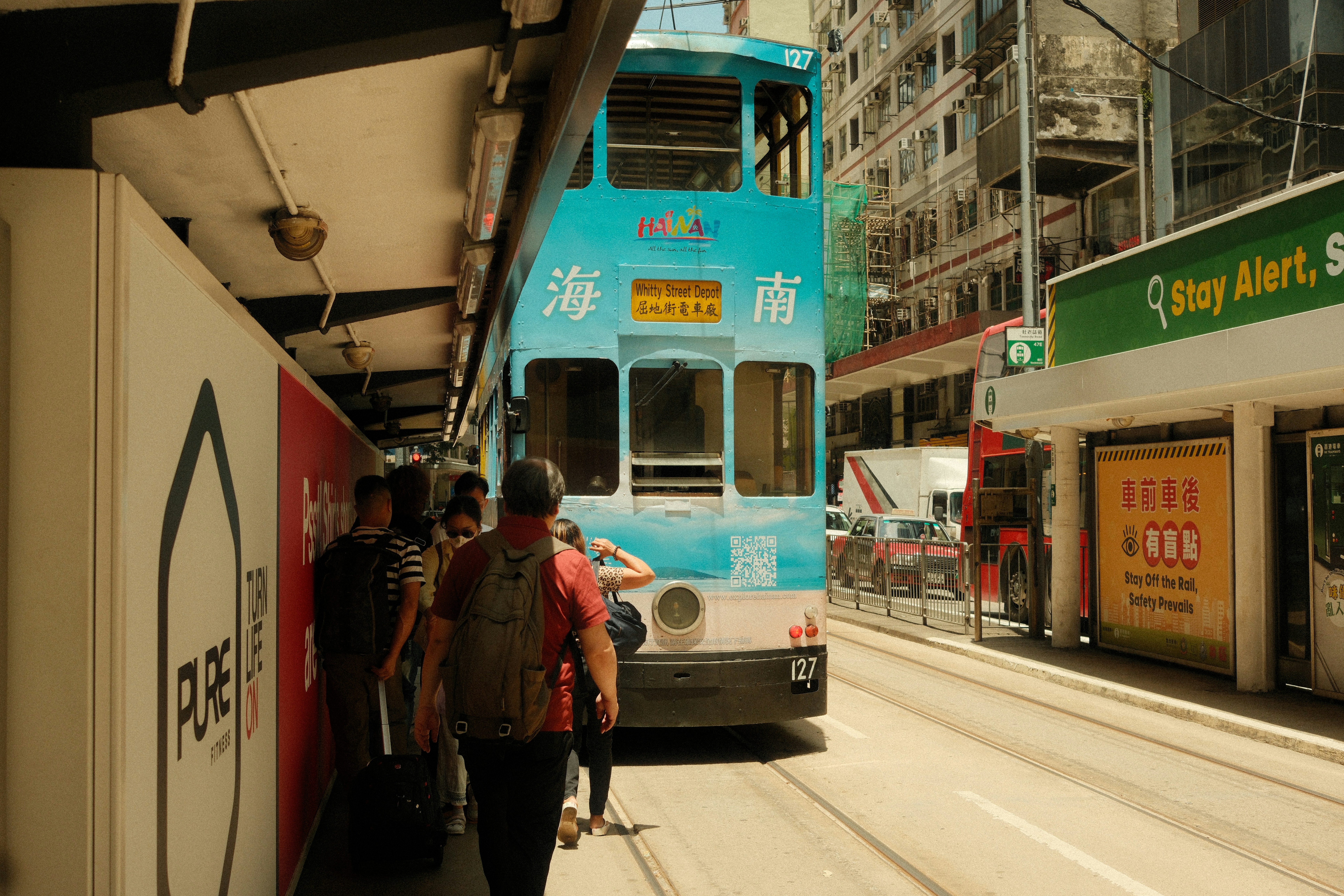 A vintage tram, adorned with vibrant blue and white colors, stands at a bustling station in Hong Kong, surrounded by commuters and city life.
