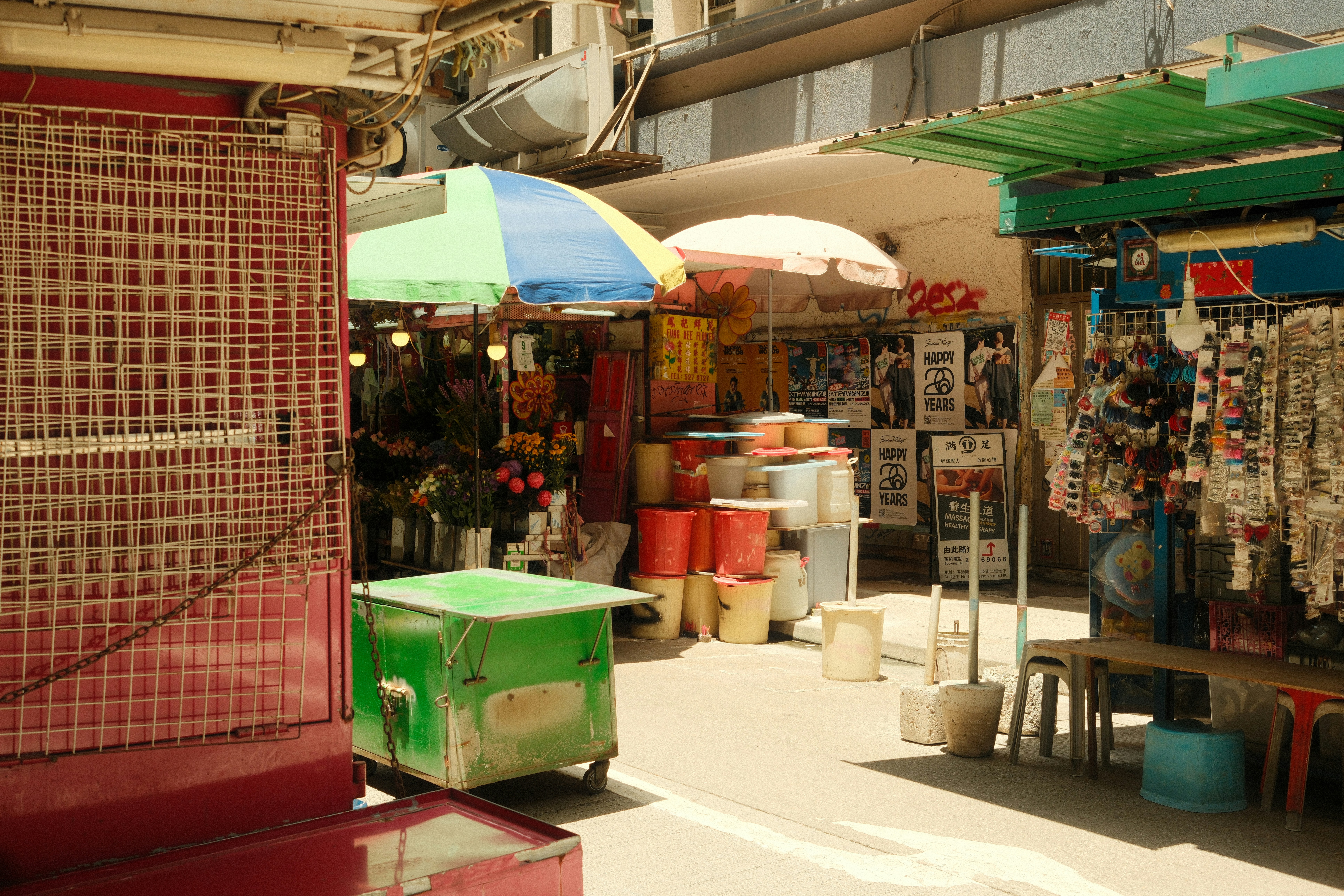 Colorful market stalls filled with flowers, buckets, and various goods create a lively street scene. Sunlight casts warm tones across the bustling atmosphere.