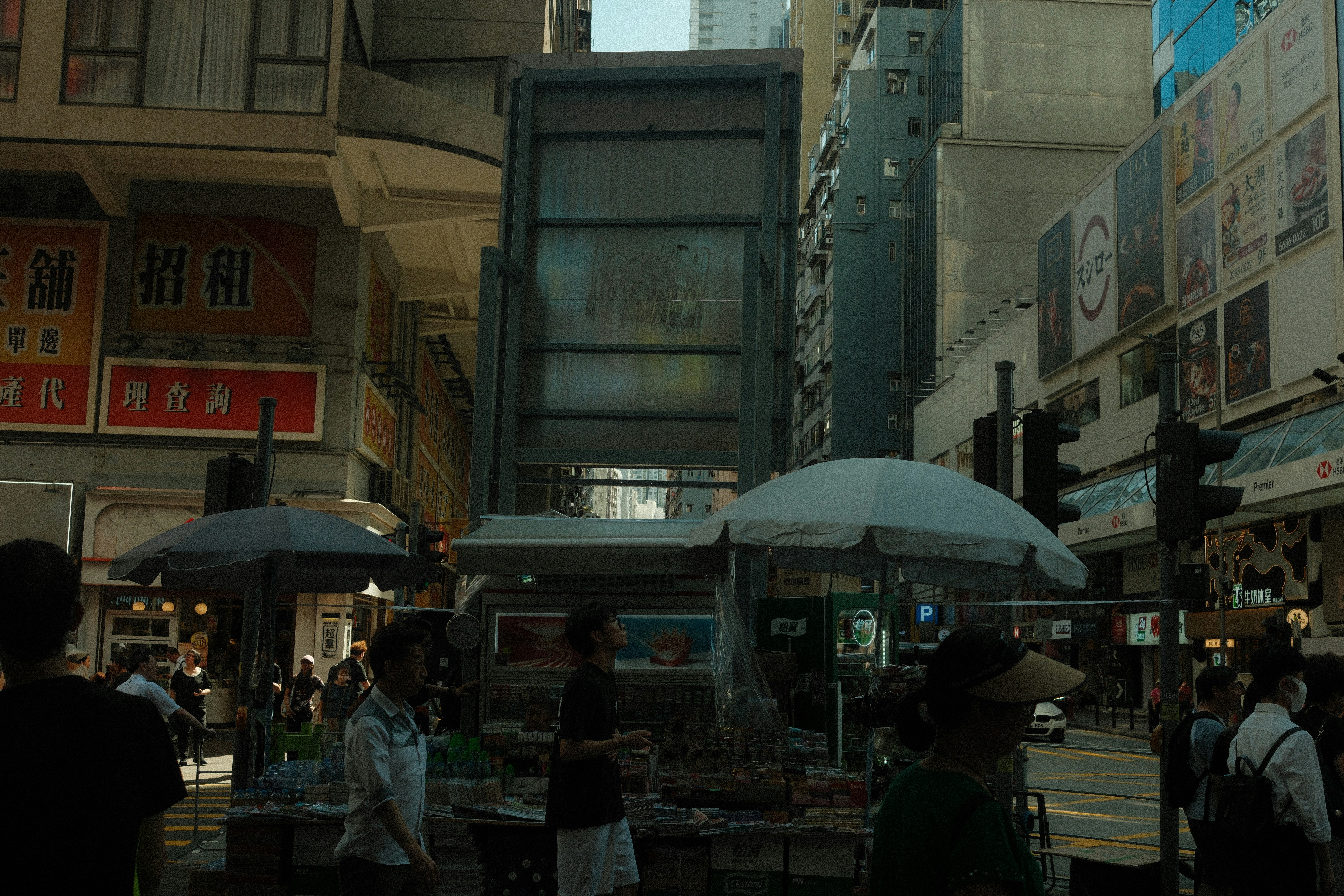 Crowded street scene in a bustling city, featuring market stalls and pedestrians navigating through vibrant storefronts. Umbrellas provide shade amidst the urban landscape.
