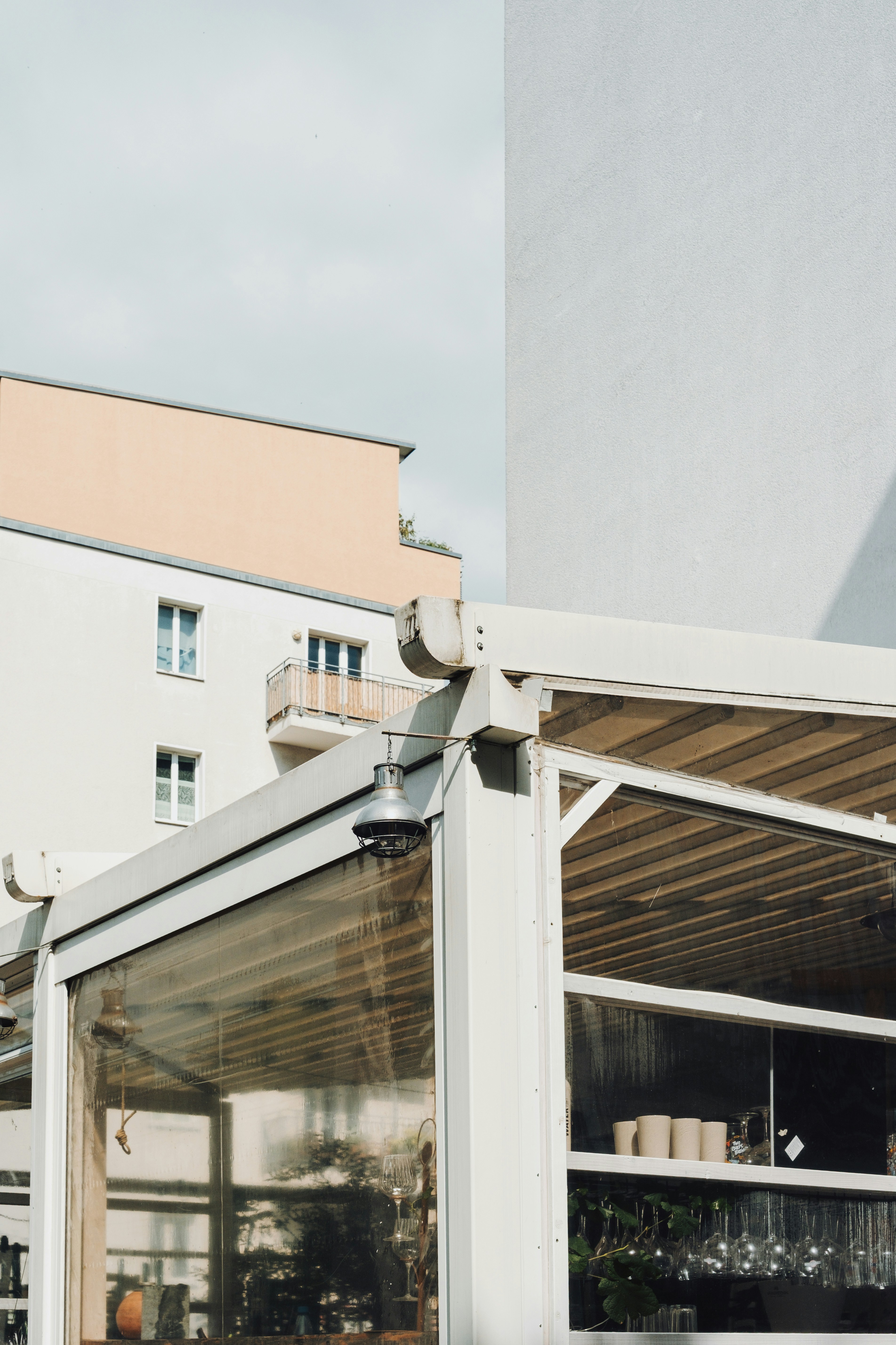 Modern glass structure with shelves of plants and utensils, set against a backdrop of urban architecture. Natural light enhances the inviting atmosphere.
