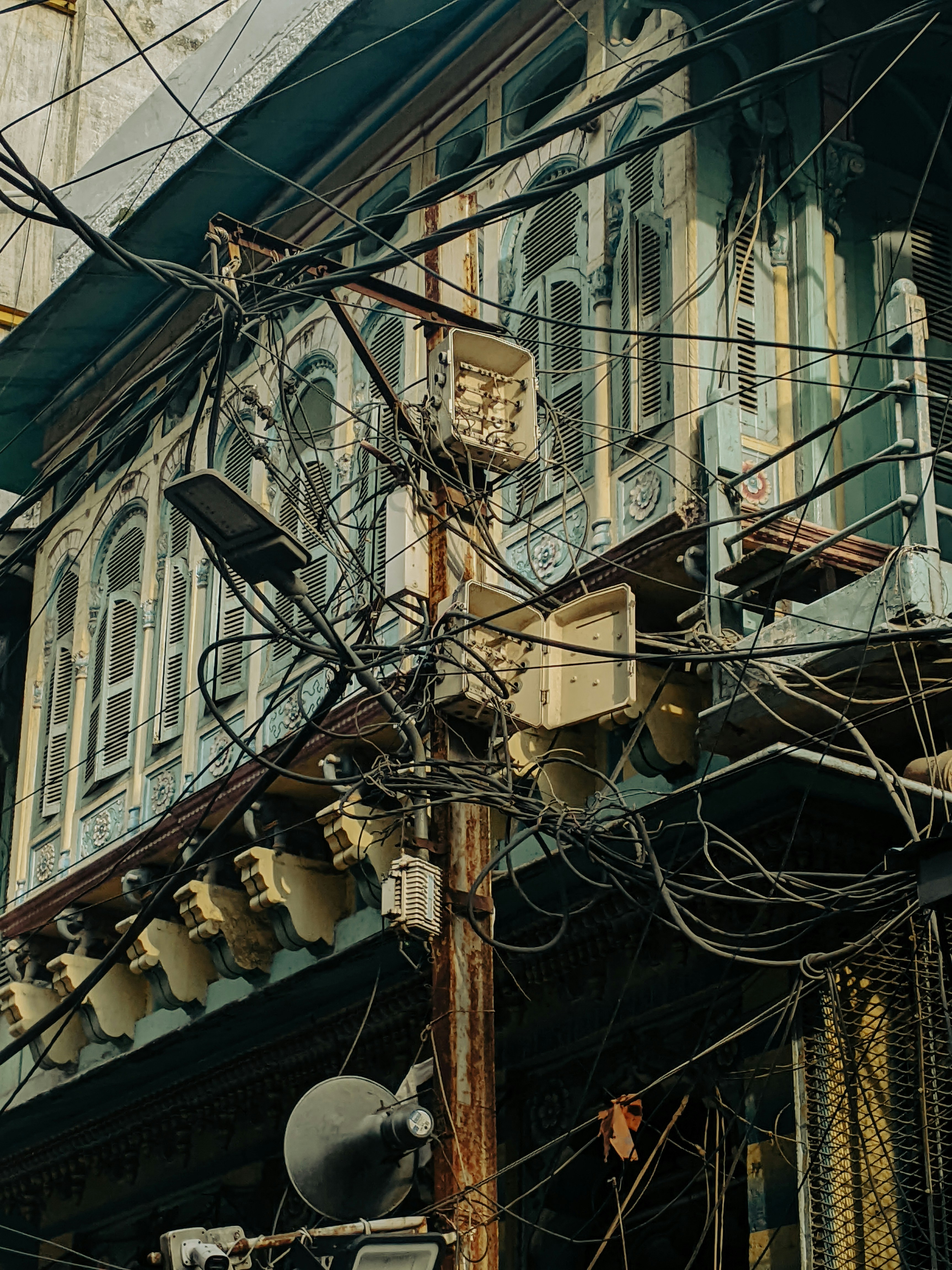 A rustic electricity pole tangled with chaotic wires, a raw slice of urban India. | Overhead wires clutter the facade of an old building.
