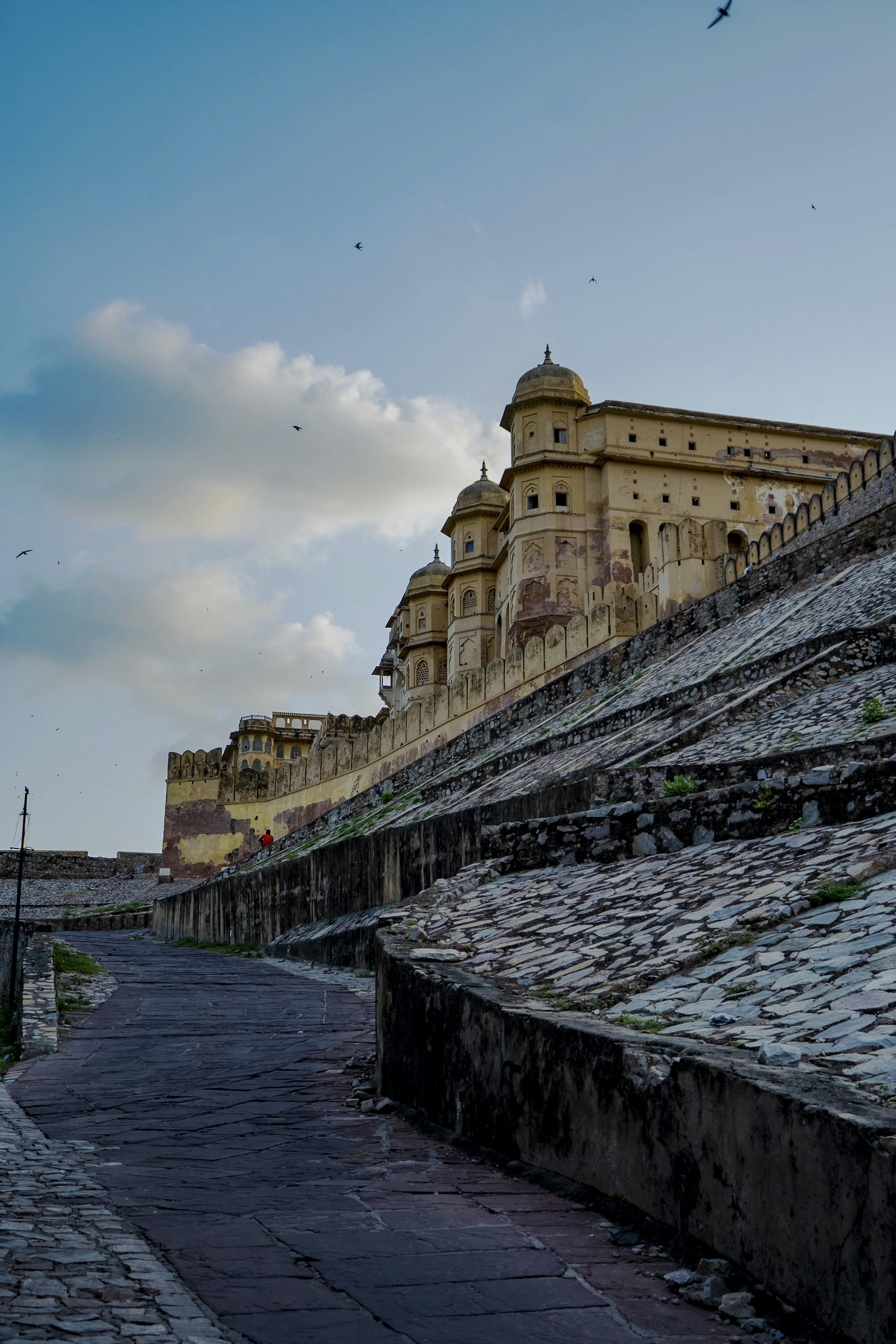 Title: Path to the Past – Amer Fort A majestic view of the historic Amer Fort in Jaipur, Rajasthan, captured from a low-angled stone pathway leading up to its grand façade. The soft evening light enhances the intricate architecture, domes, and weathered textures of this 16th-century fortress. Birds soar above, adding motion to the stillness of this iconic heritage site — a perfect blend of history, scale, and serenity | An ancient indian fort stands tall against the sky.