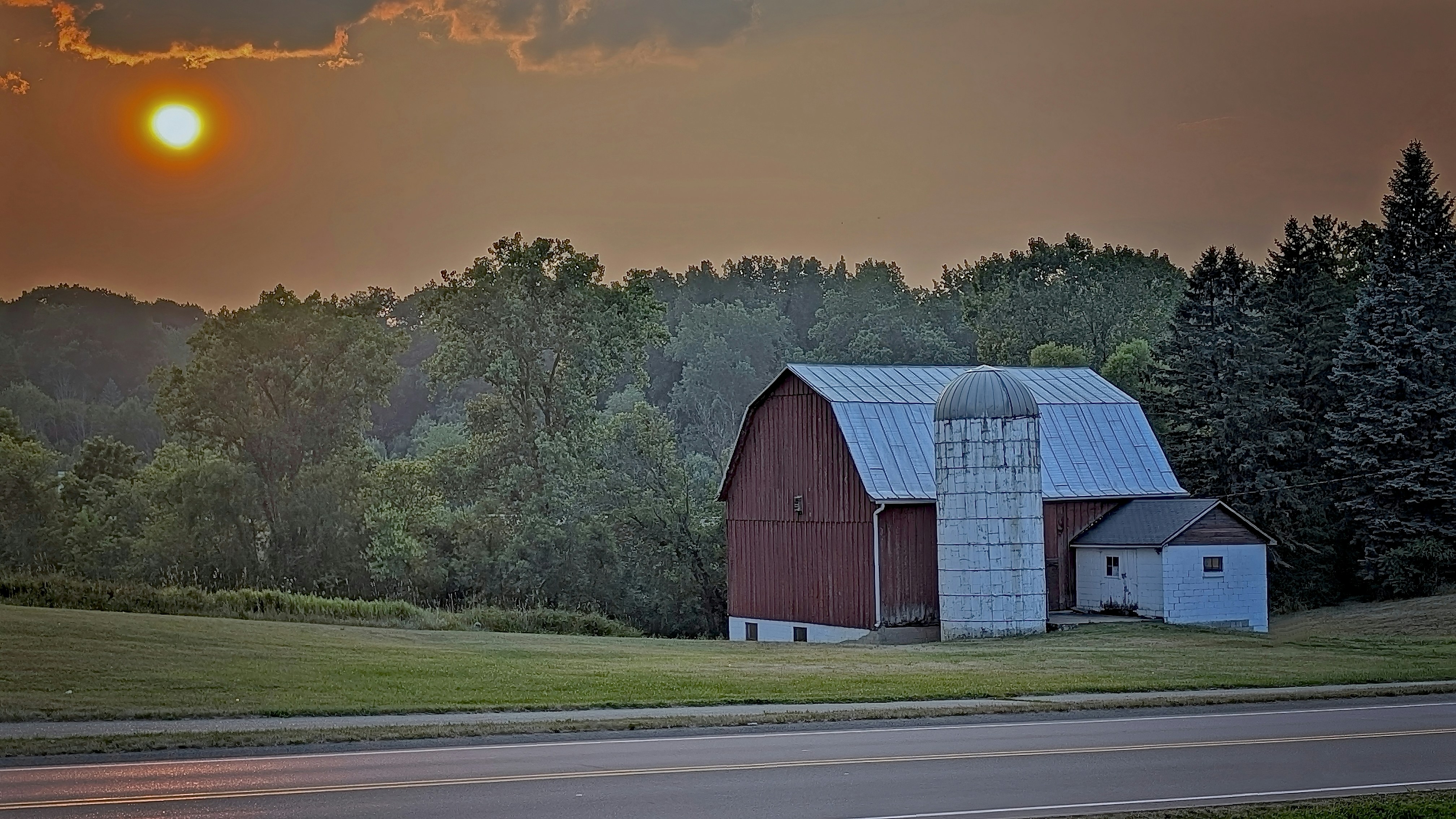 Red barn with a silo nestled among lush greenery under a setting sun, evoking a sense of tranquility.