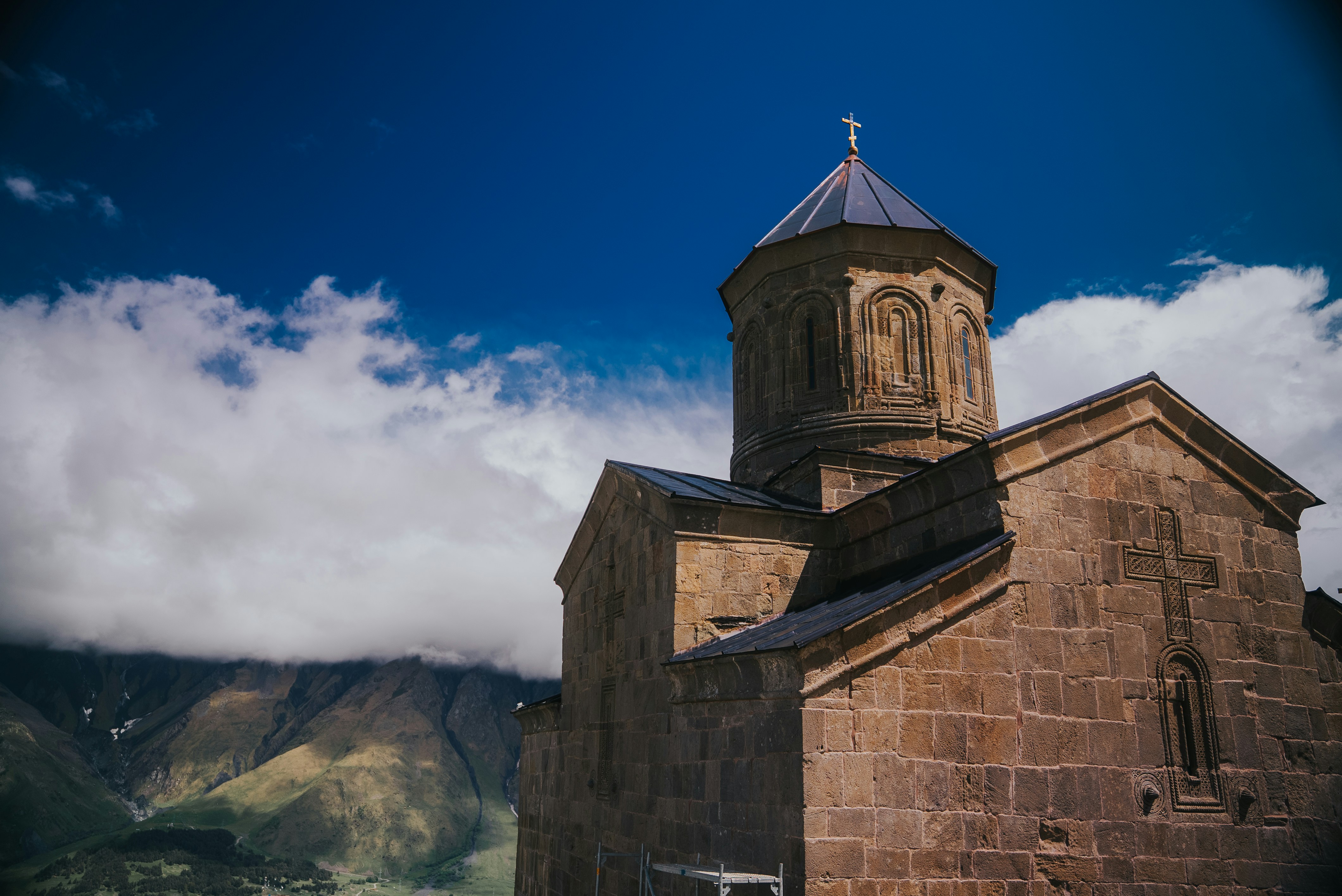 Historic church nestled in the mountains, showcasing intricate stonework under a dramatic sky. Clouds loom in the background, adding depth to the serene landscape.