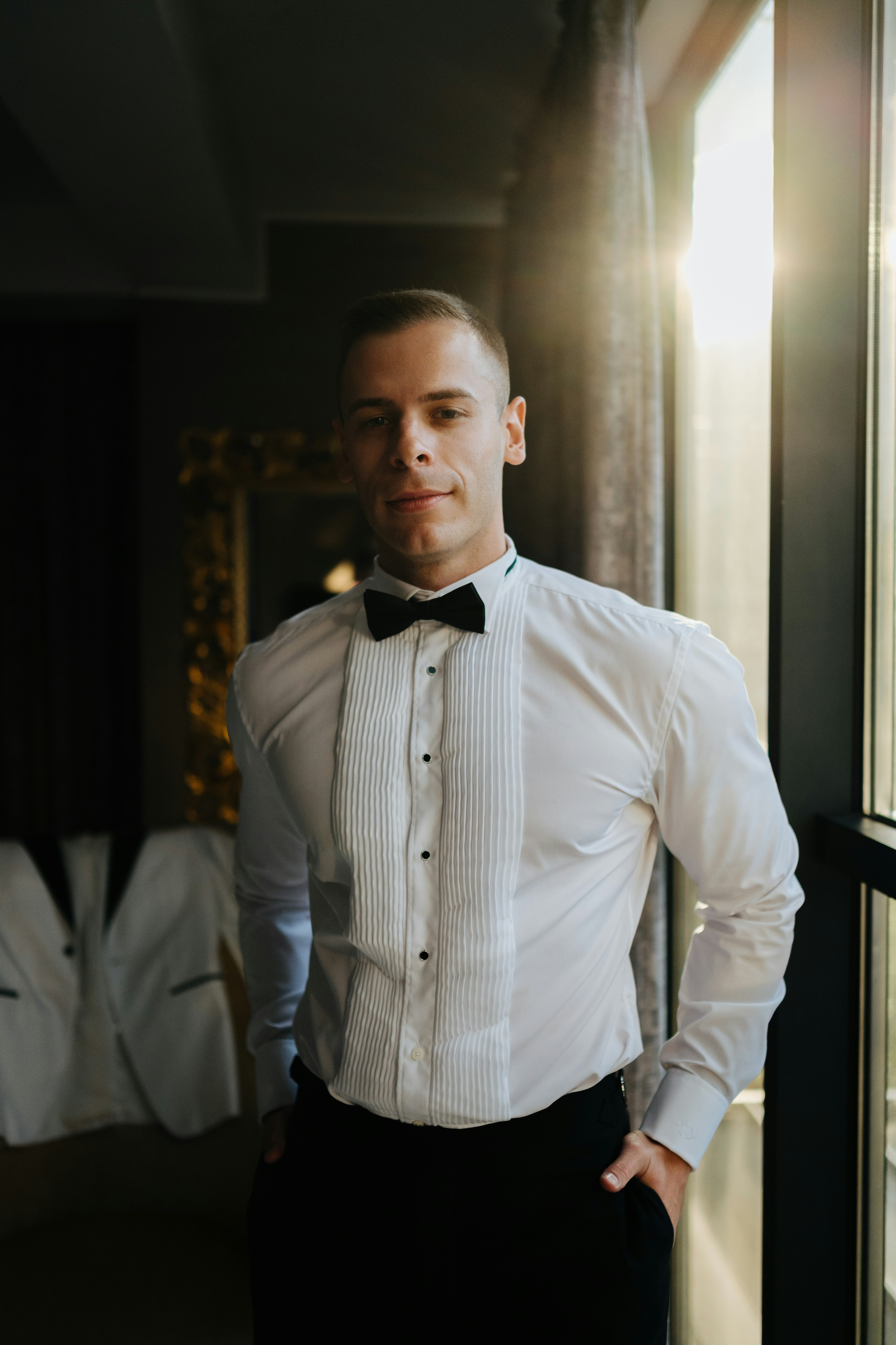 A man in a tuxedo poses by a window.