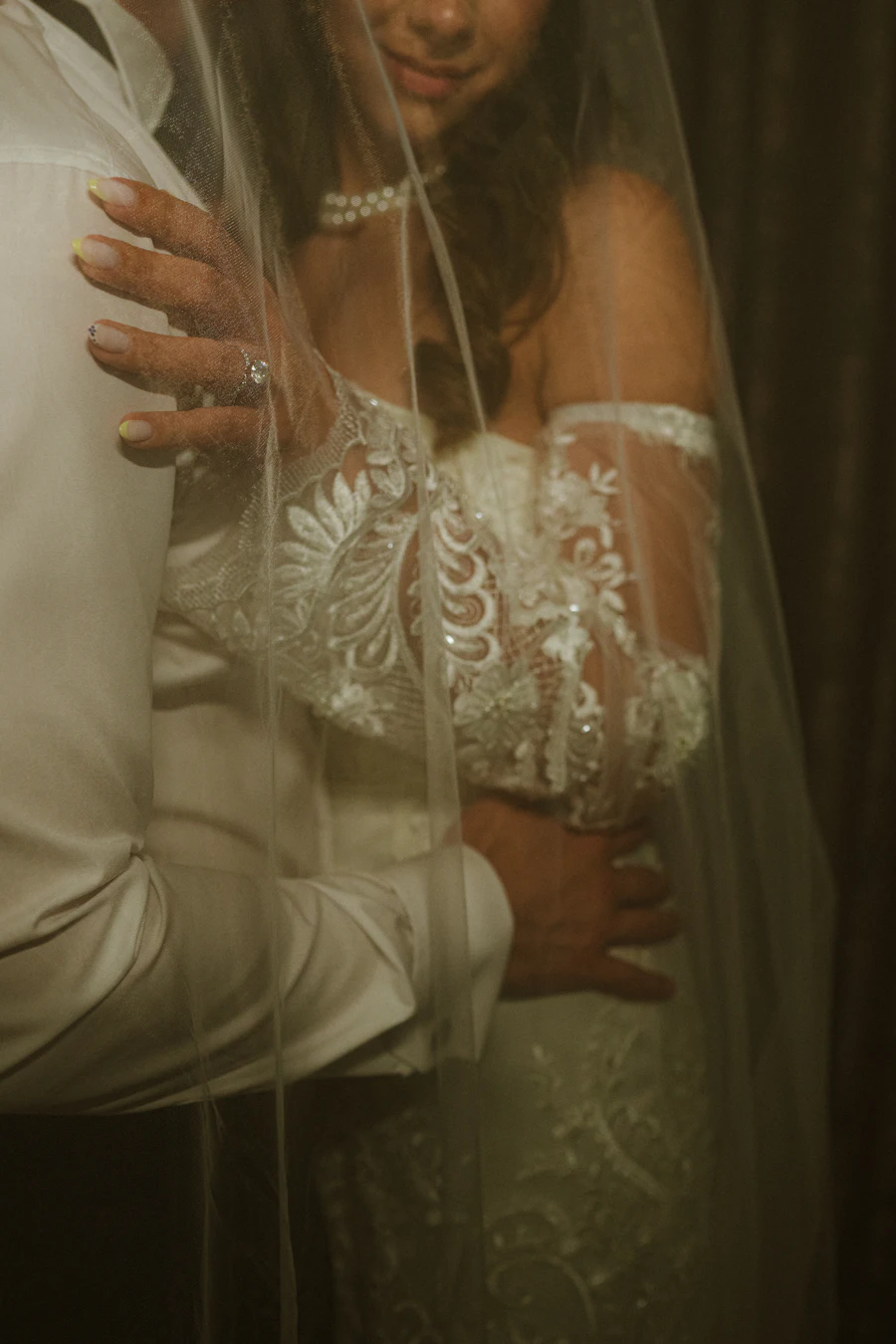 Bride in an embroidered off-shoulder gown and veil being embraced, with a hand on her shoulder and another at her waist. Soft, warm light gives the scene a dreamy, intimate feel.