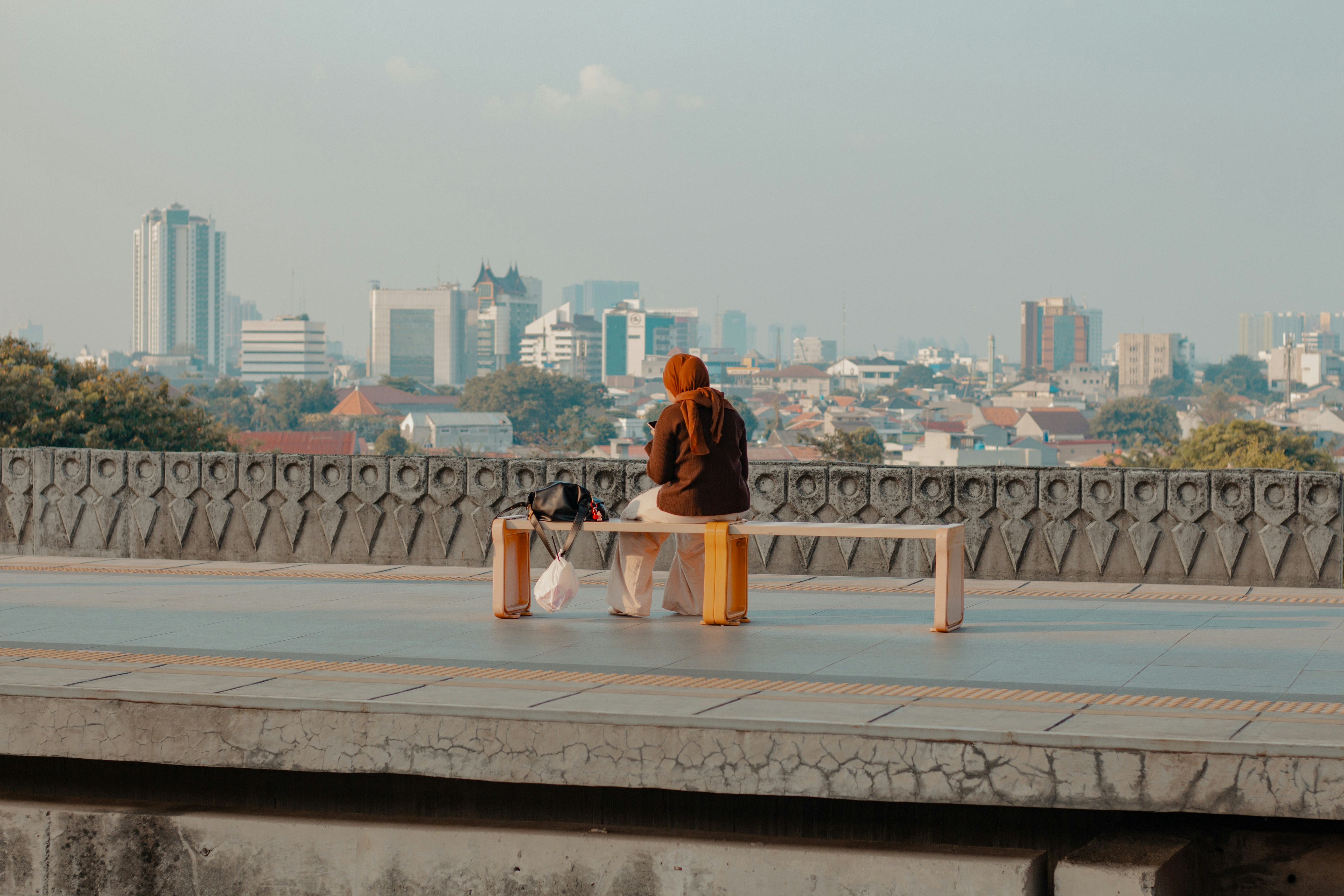 Riverside Solitude: A Hooded Figure Benchside Against the Distant Cityscape