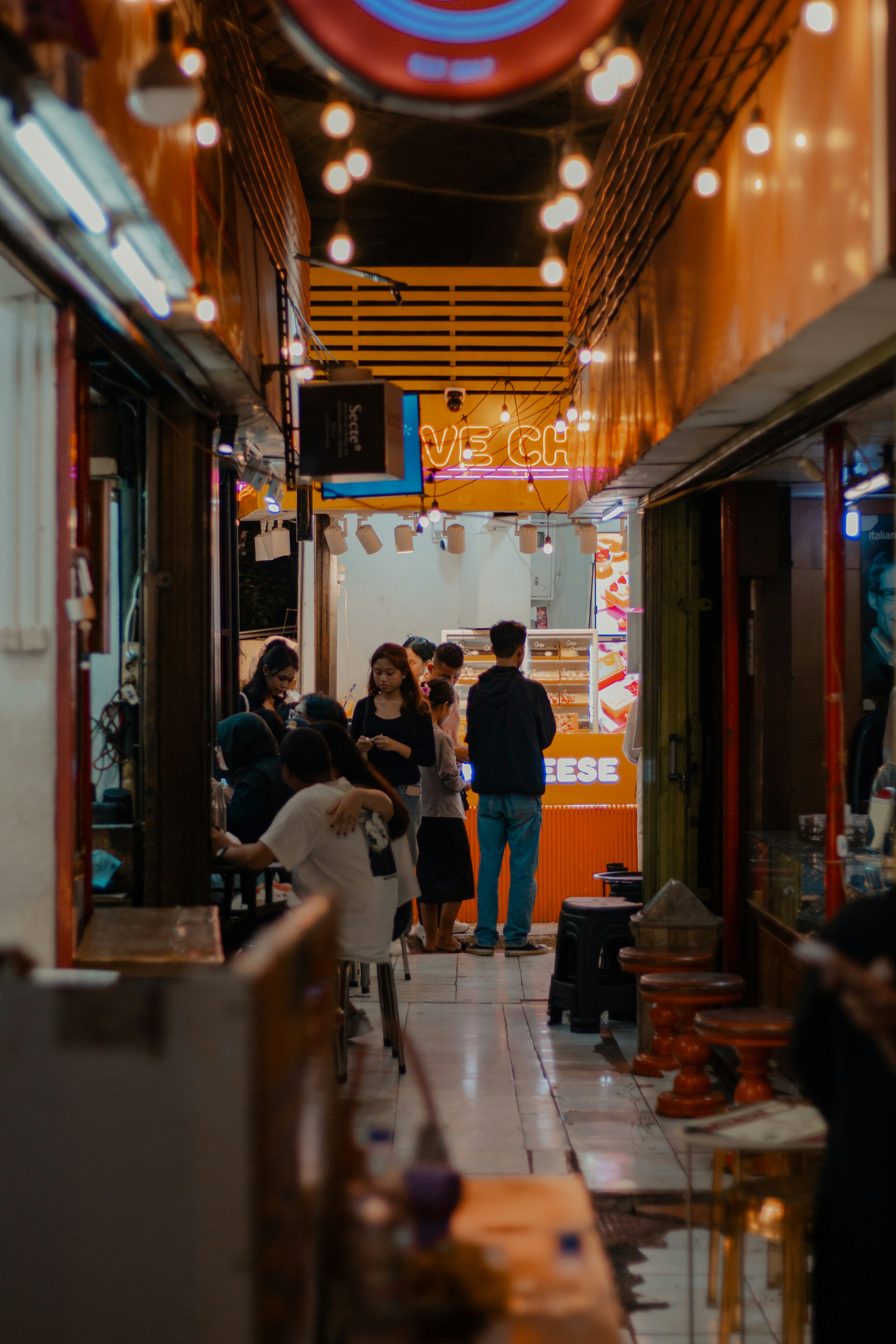 Vibrant alleyway bustling with people, illuminated by colorful neon signs and warm string lights, showcasing a lively food scene.
