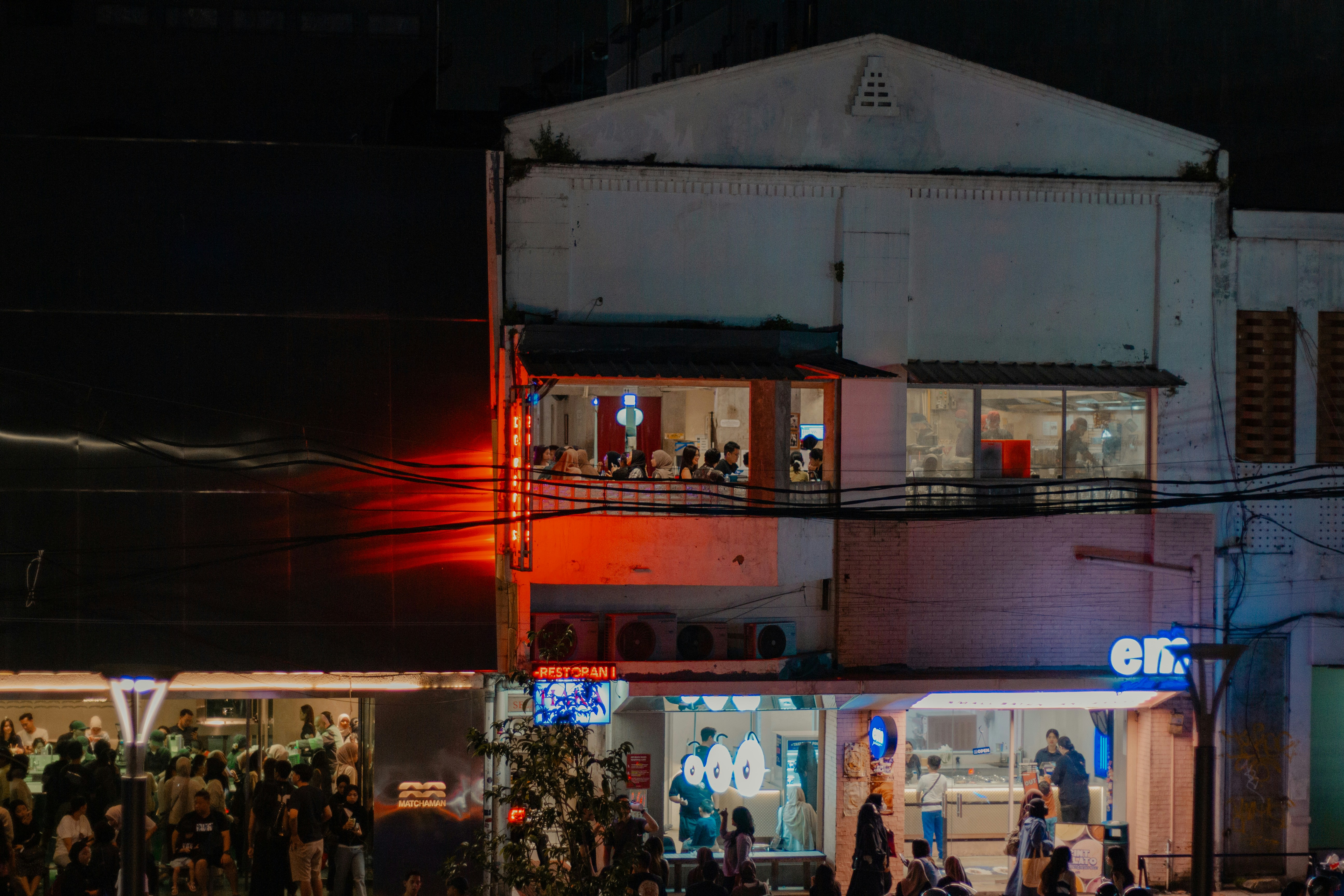 A vibrant street scene showcasing a bustling eatery with patrons enjoying their meals, illuminated by colorful lights. The architecture reflects a blend of modern and traditional styles.