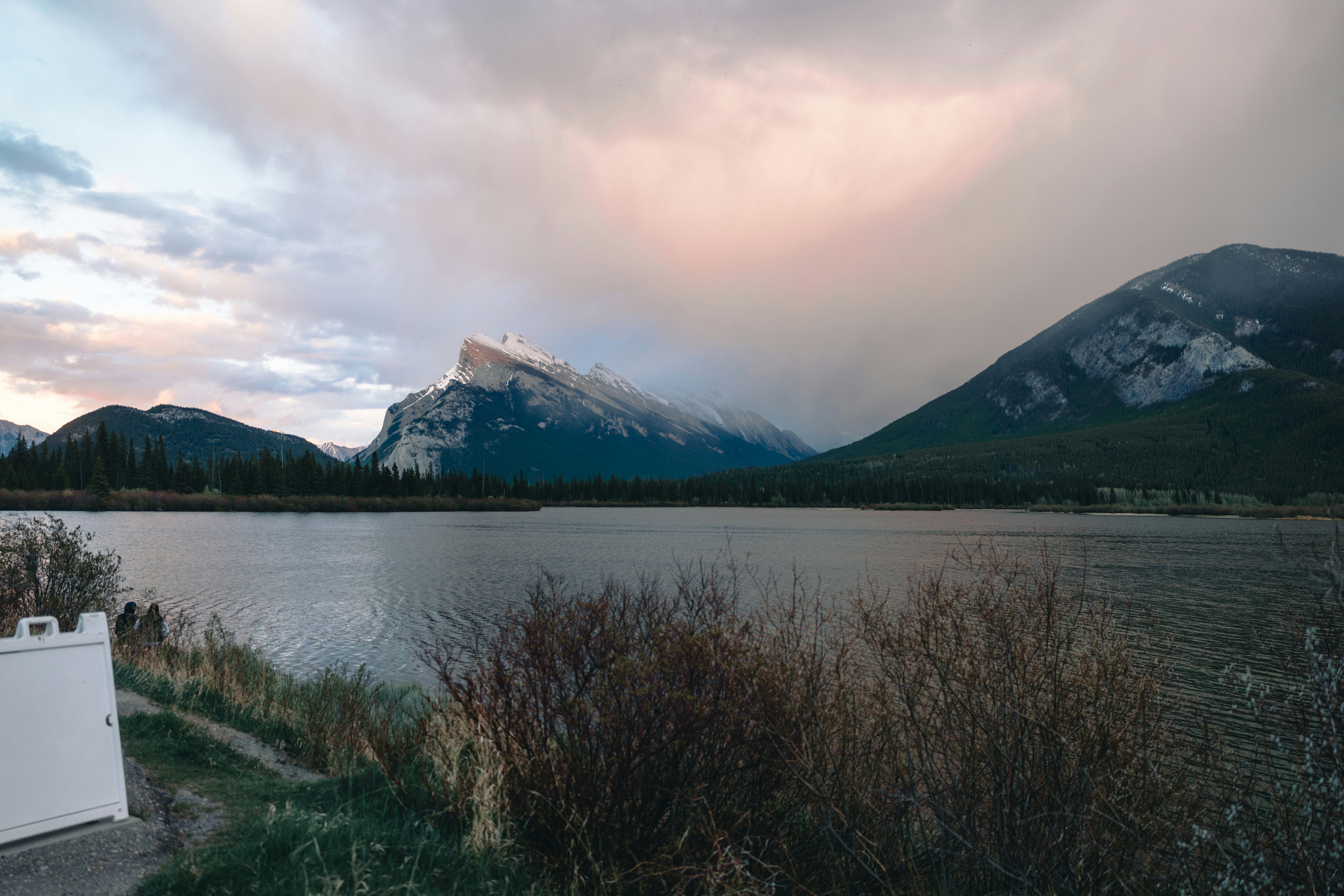 A tranquil lakeside view featuring majestic mountains under a moody sky, with hints of sunlight illuminating the peaks. The foreground includes soft vegetation and a pathway leading to the water's edge.