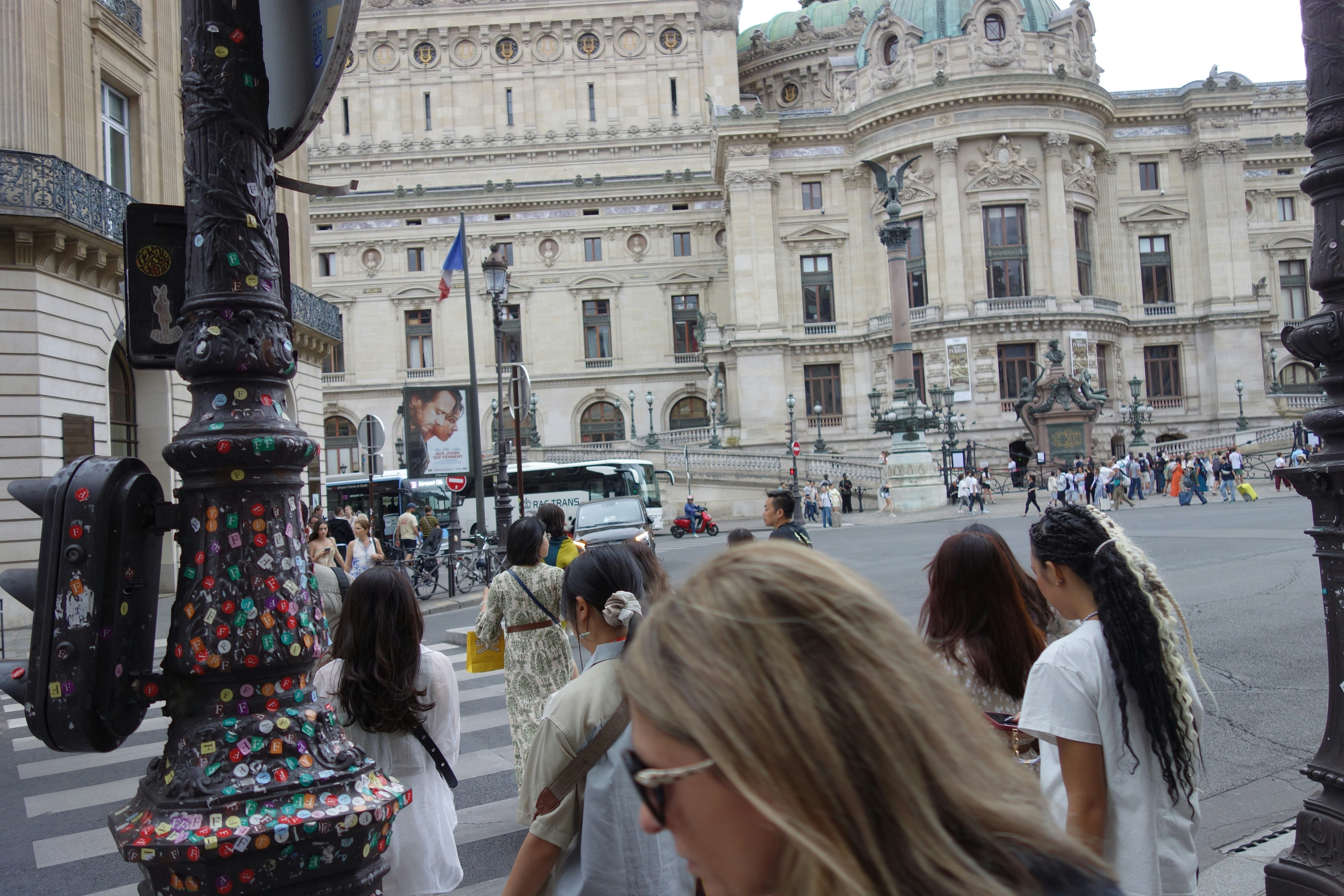 A bustling street scene showcasing pedestrians crossing near an ornate building, with a colorful, sticker-adorned lamppost in the foreground.