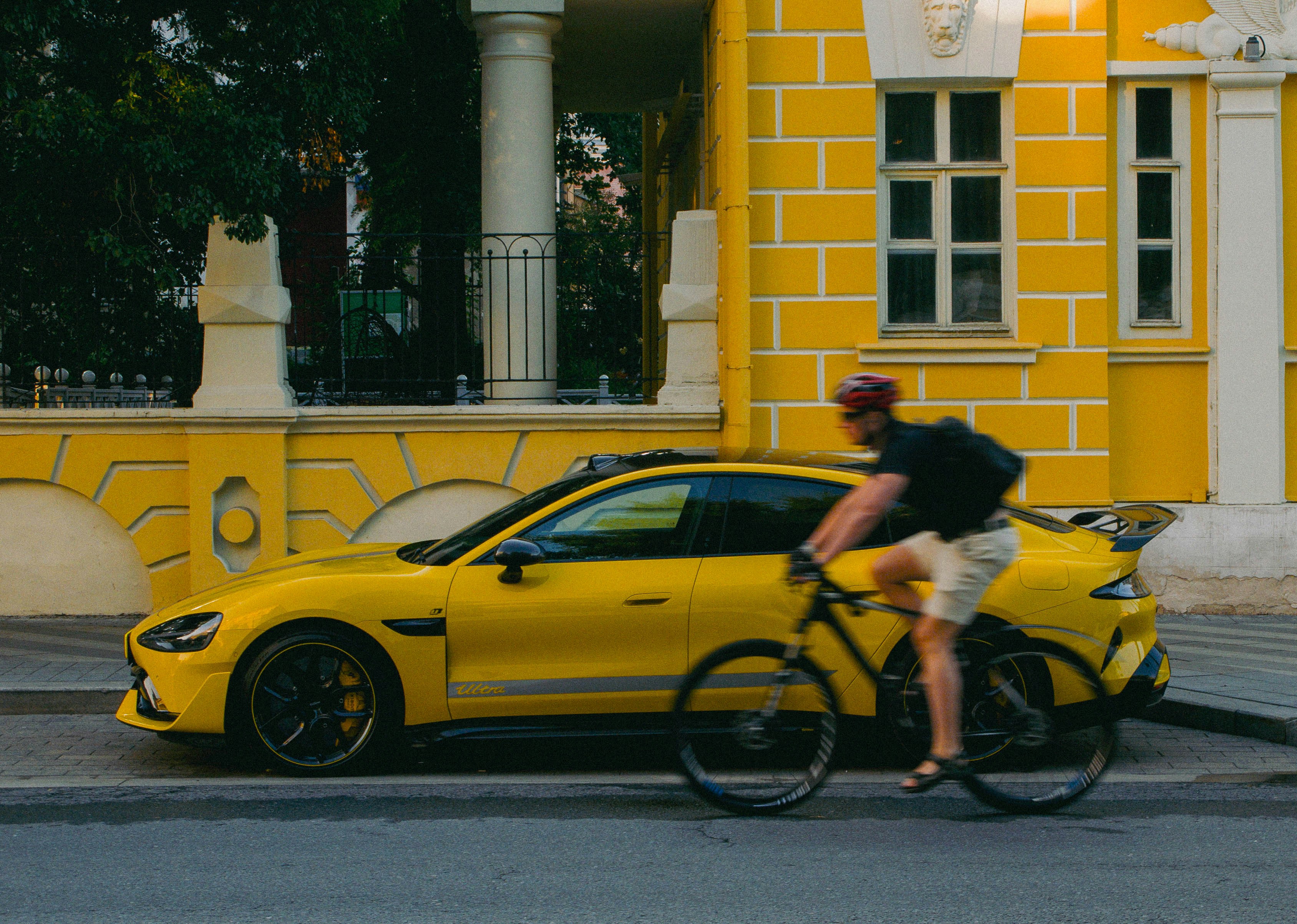 A cyclist rides past a yellow car and building.
