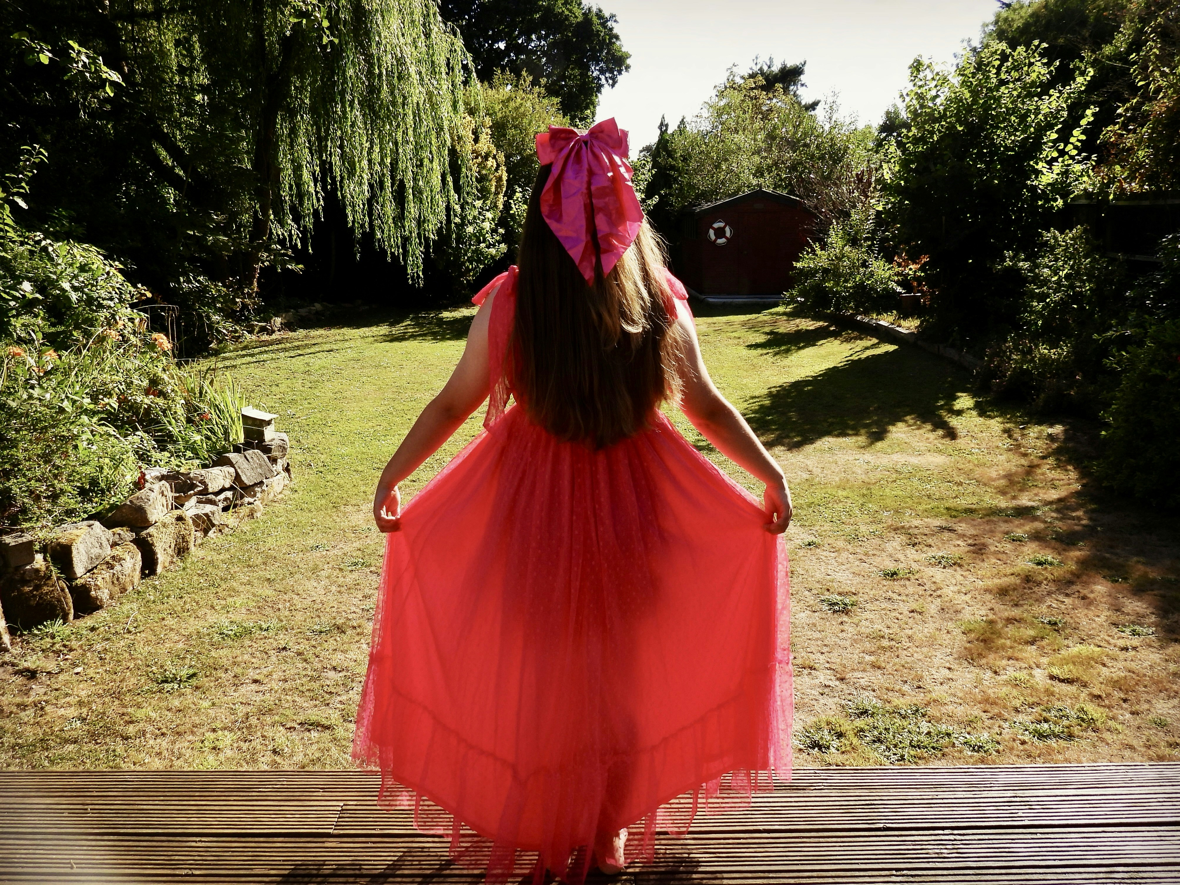 A young girl in a vibrant pink dress and large bow stands with her back to the viewer, surrounded by a lush garden setting. The sunlight casts a warm glow over the scene.