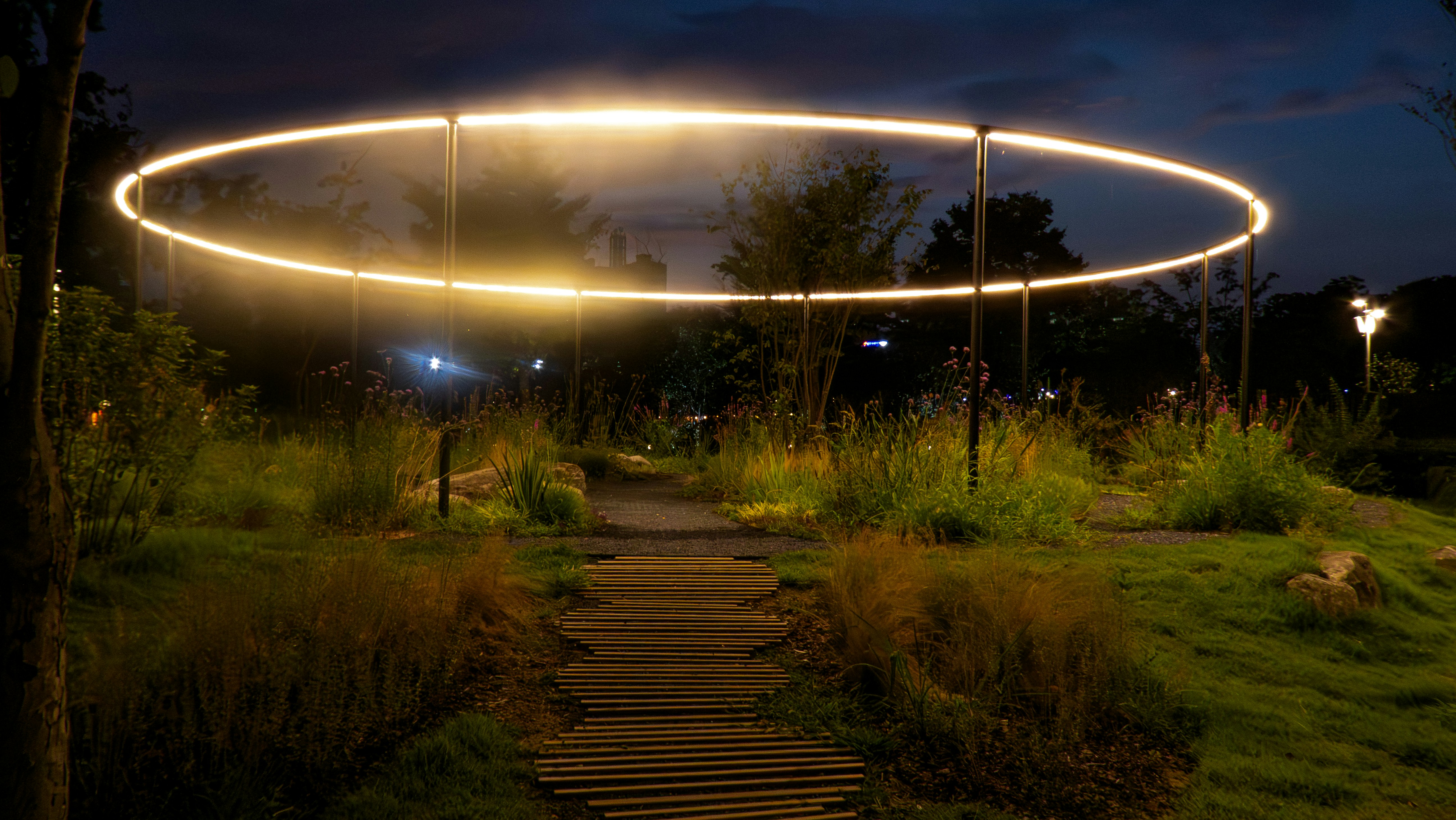 Circular light installation glowing softly above a lush garden path, framed by vibrant greenery and twilight hues.