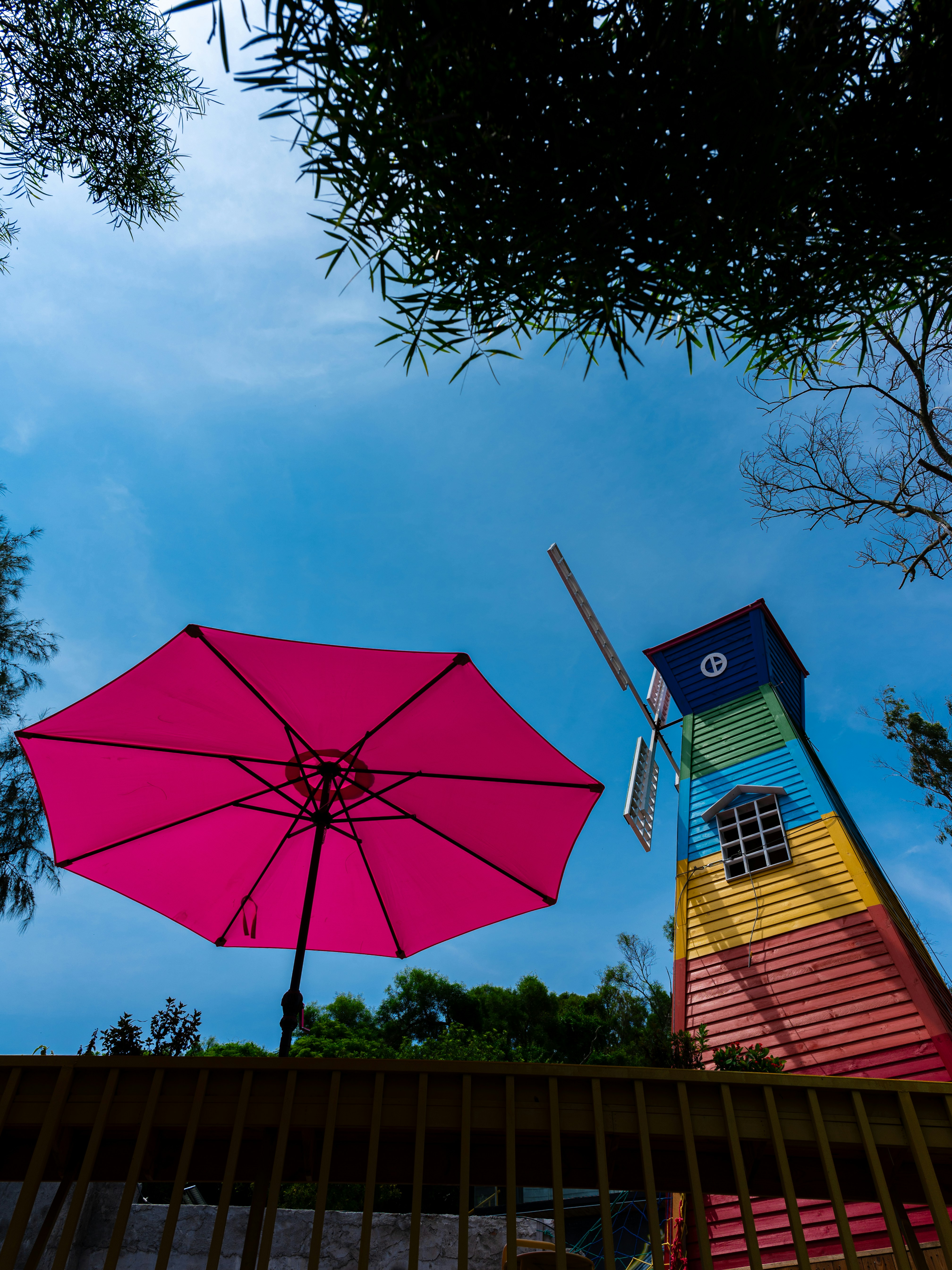 Vibrant pink umbrella stands tall against a colorful windmill and bright blue sky. The scene captures a playful blend of nature and whimsical architecture.