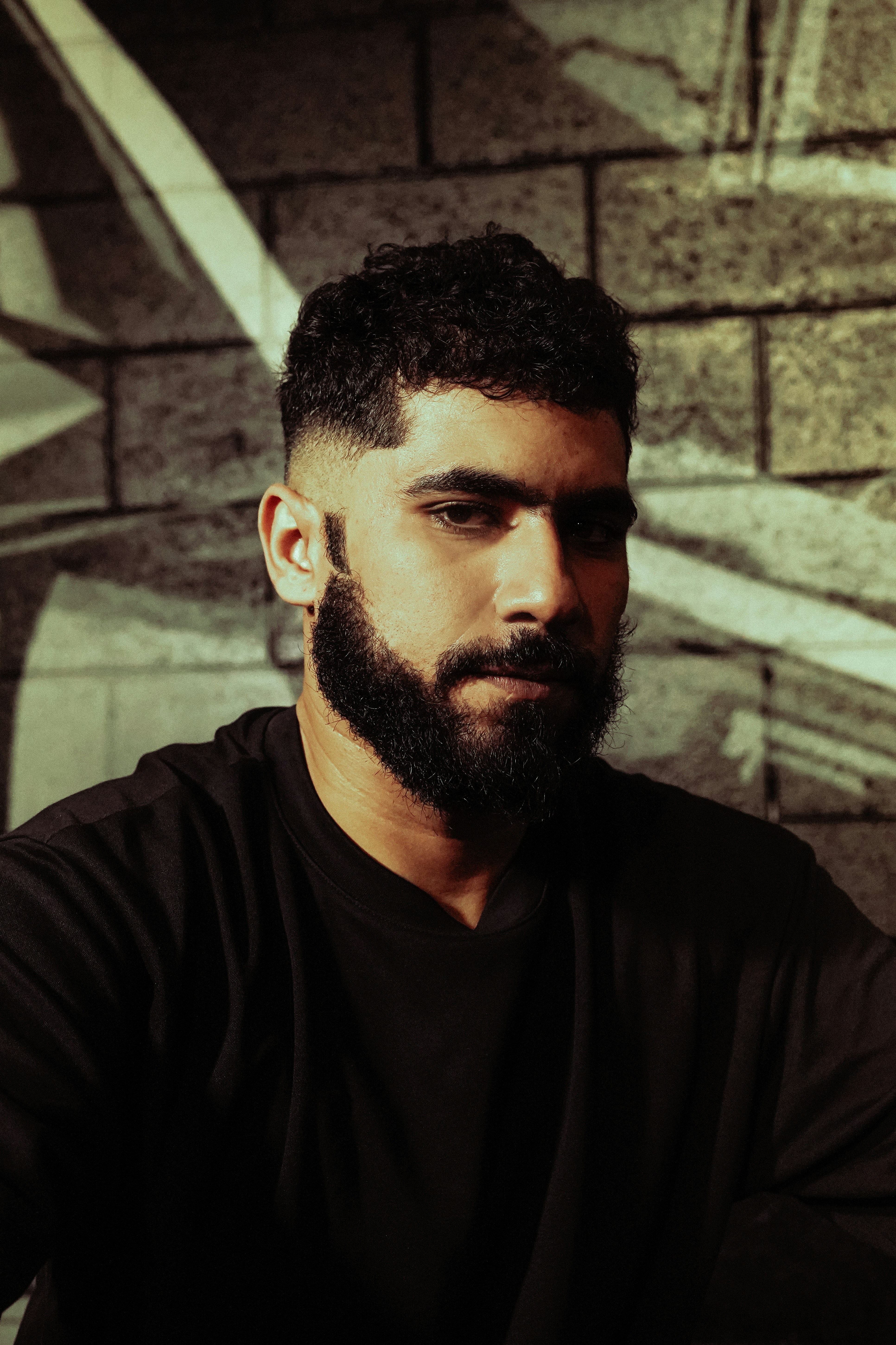 A man with a thick beard and short hair poses against a textured wall, illuminated by soft lighting that highlights his features.