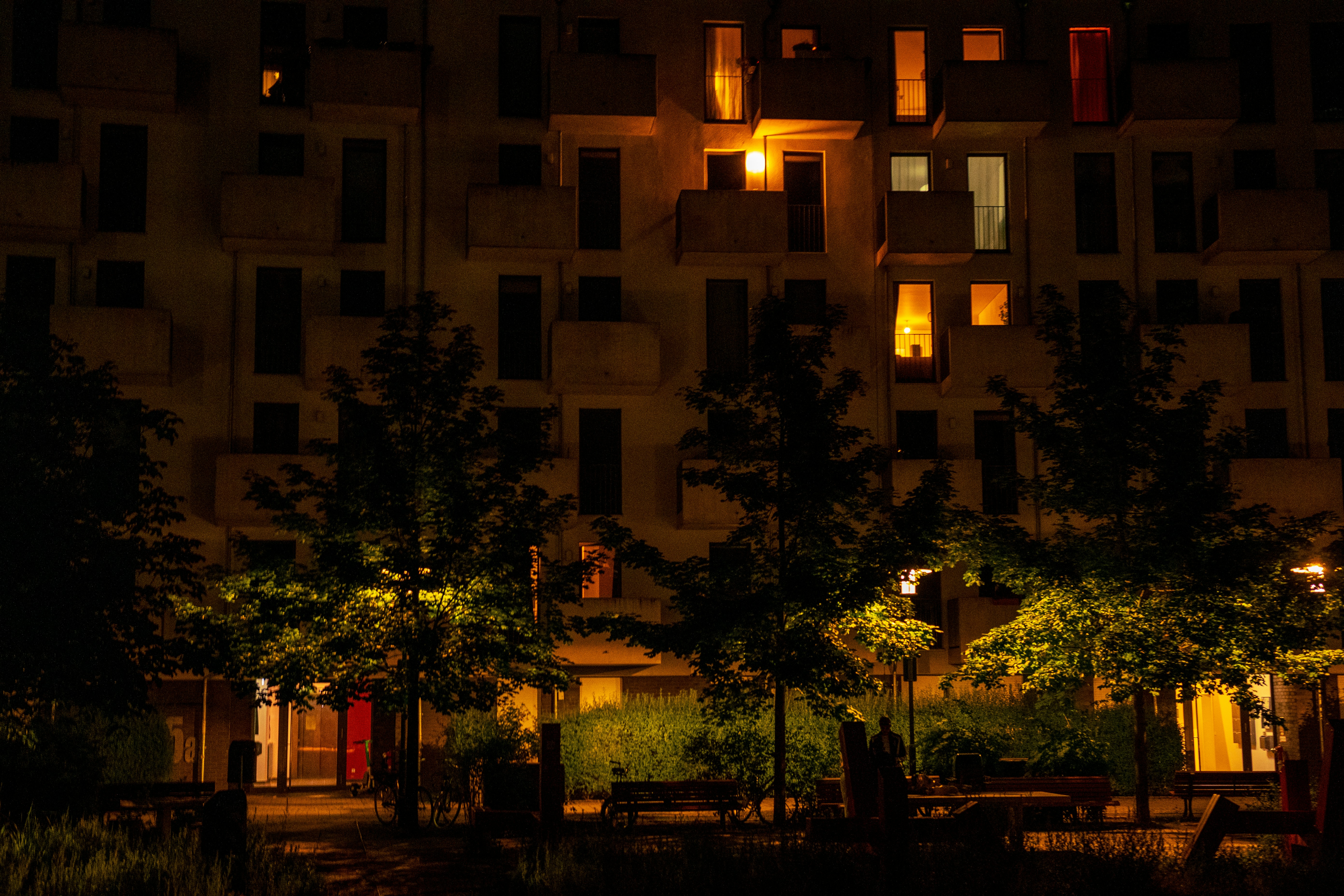 Illuminated windows of a modern apartment building reveal glimpses of life within, framed by lush greenery in a nighttime setting.