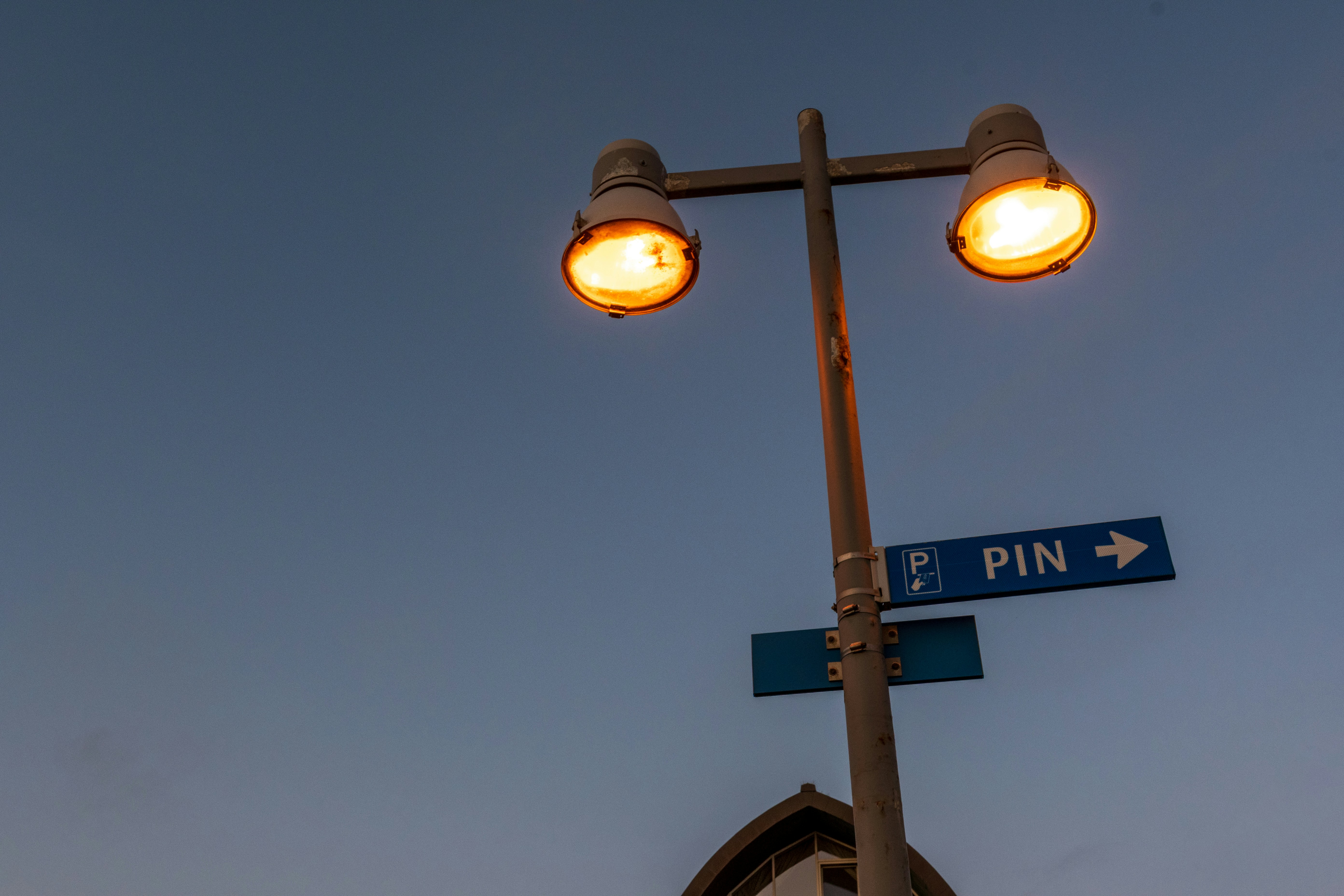 Street lamp casting a warm glow over a directional sign pointing towards parking. The scene captures the transition from day to night.