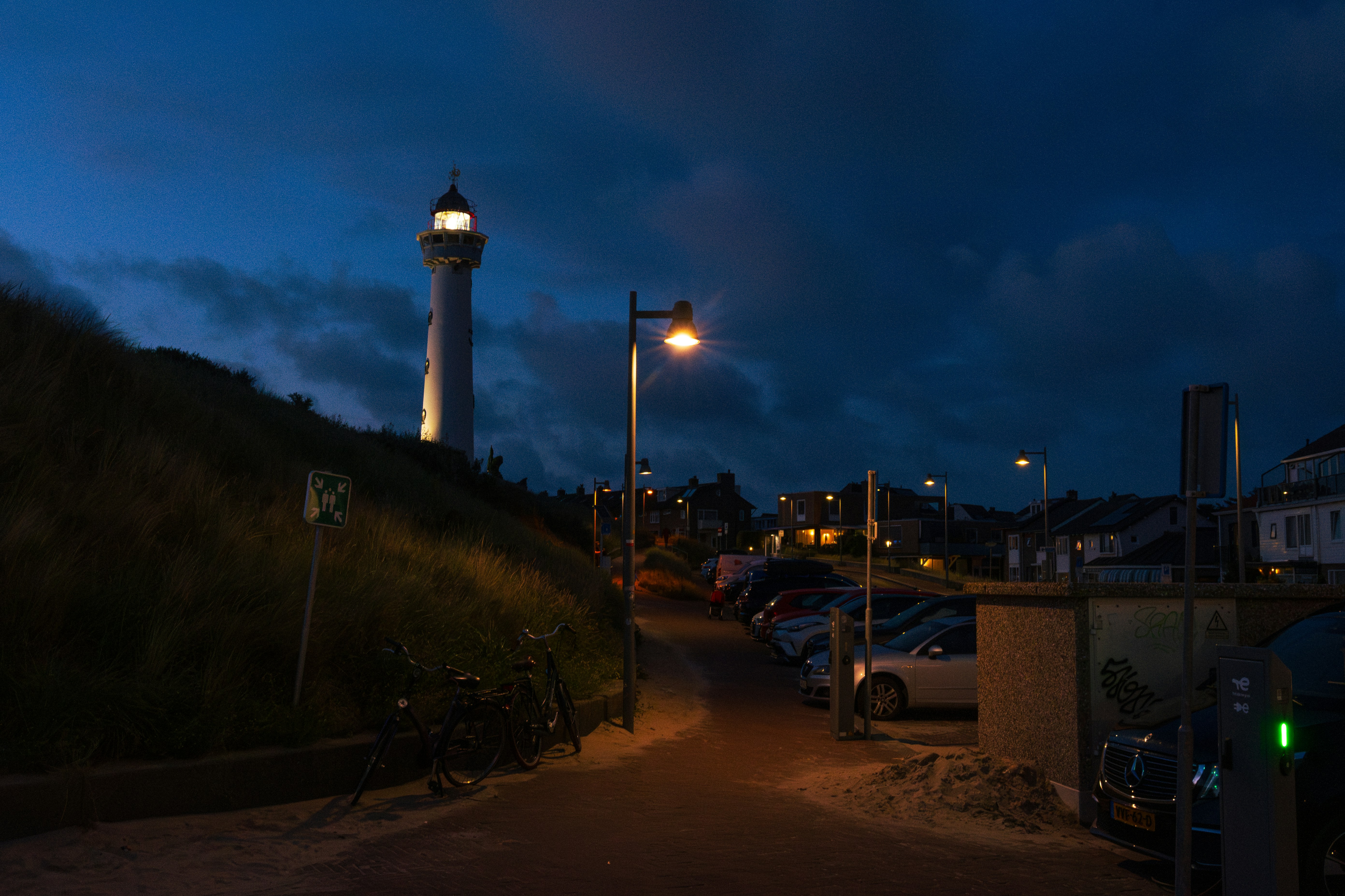 A lighthouse stands tall against a twilight sky, illuminating a coastal pathway lined with parked cars and gentle dunes.