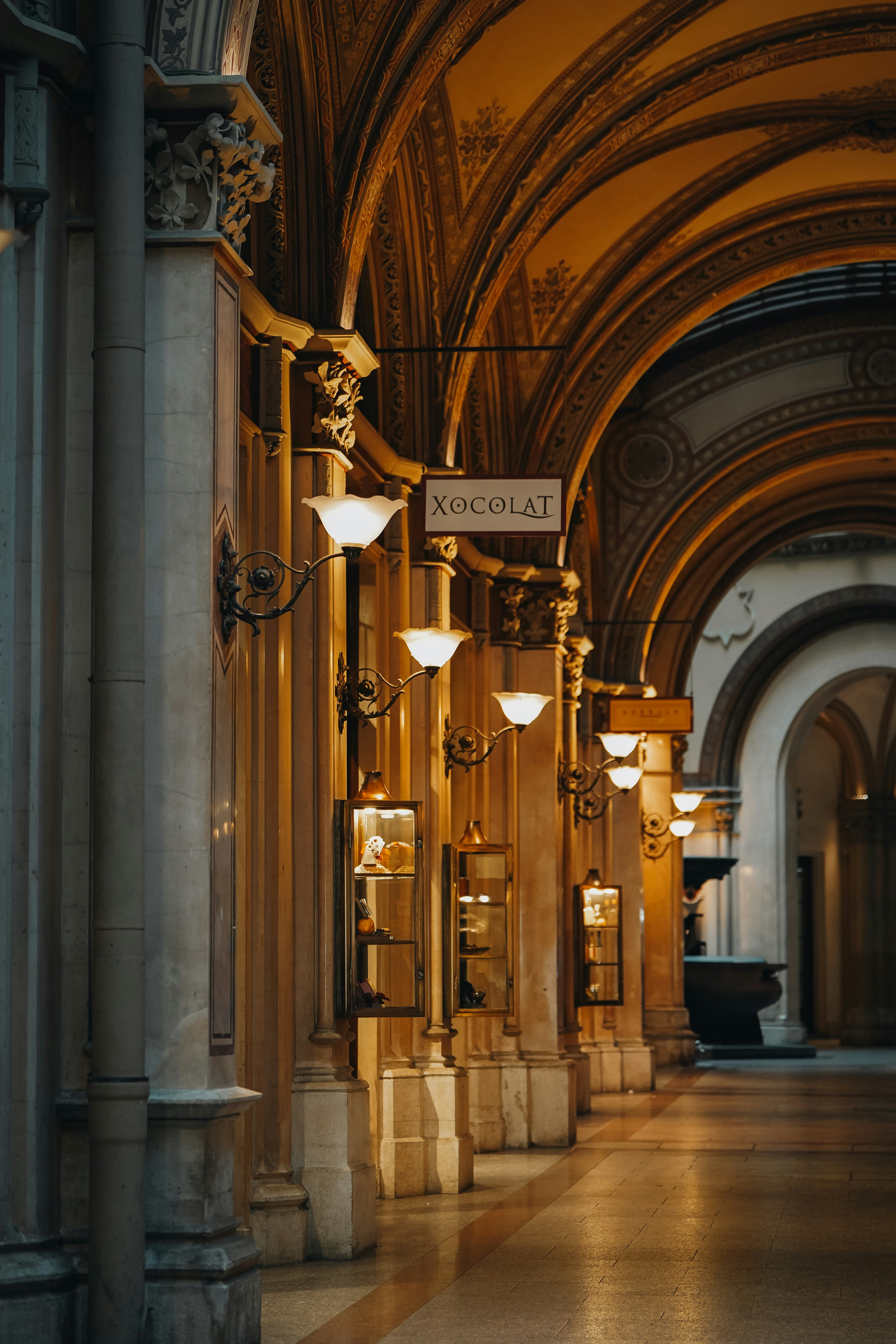 Warm lamplight spills across ornate archways, drawing you into the quiet grandeur of Vienna’s hidden arcades—refined, golden, and inviting. | A grand, illuminated hallway of a chocolate shop.