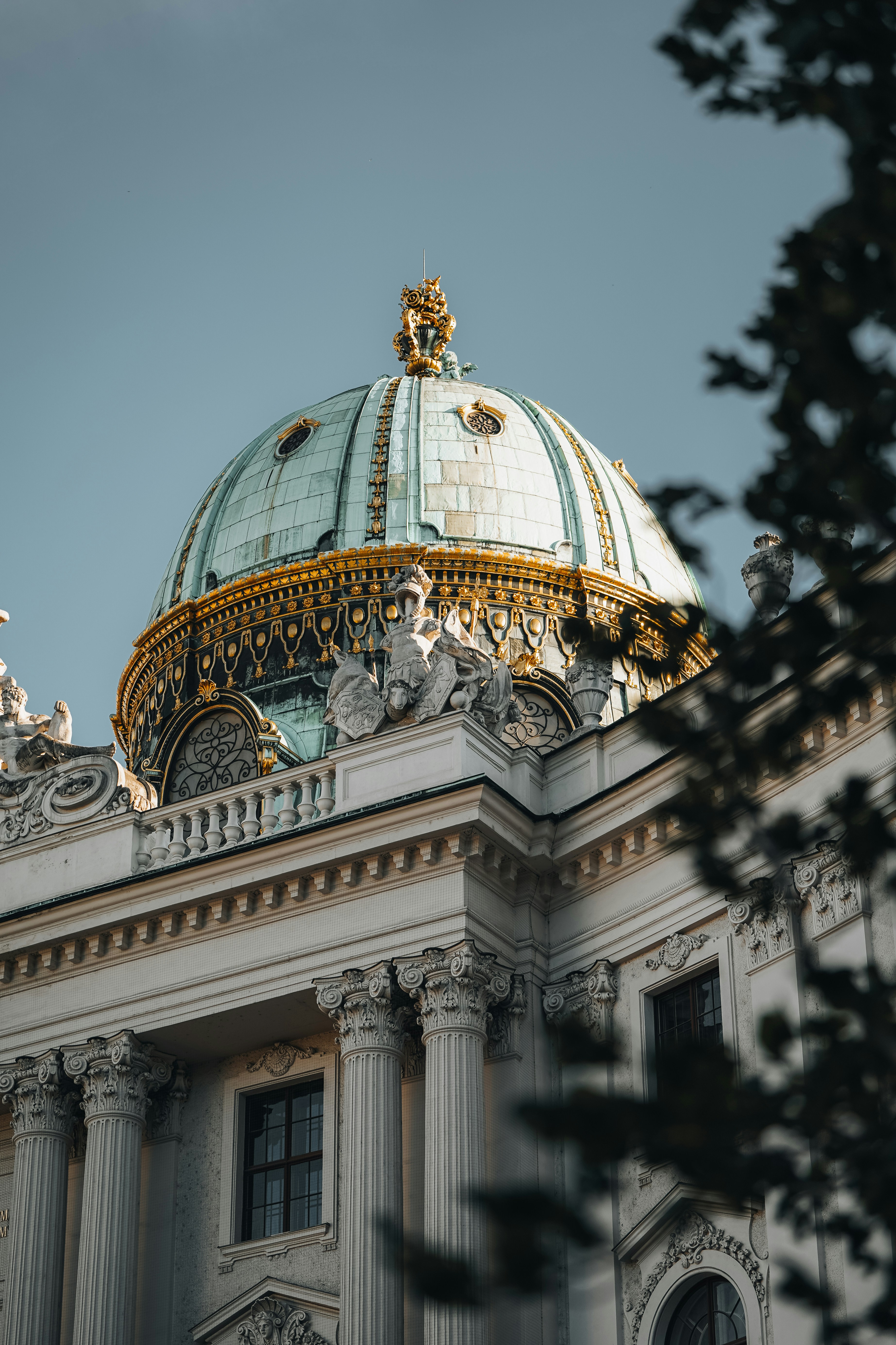 The morning light graces the baroque dome of Hofburg Palace, where every detail—from golden flourishes to the watchful sculptures—tells a story of imperial elegance. | Ornate building dome against a clear sky.