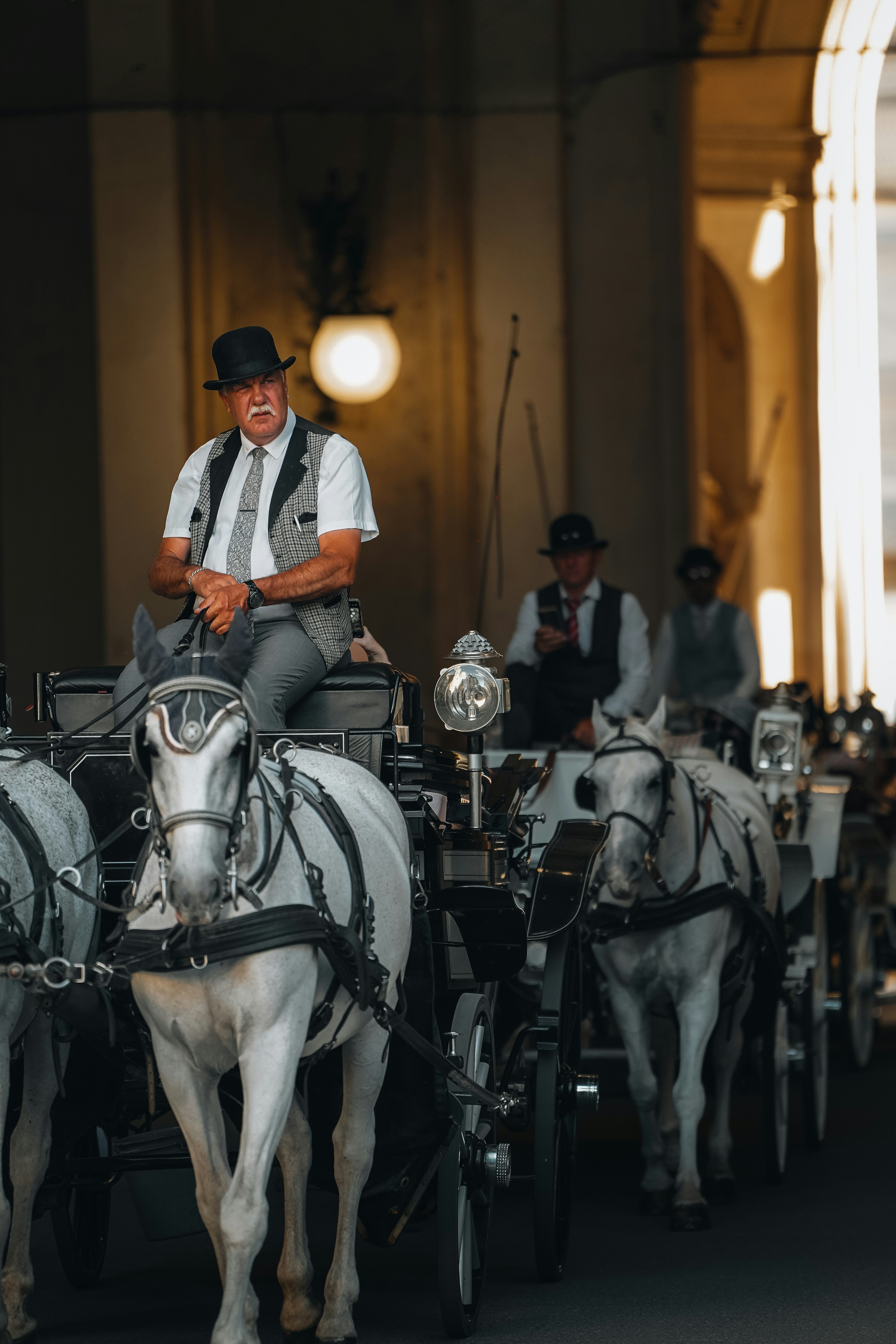 Tradition rides through shadowed arches as a Fiaker driver steers his white horses—Vienna’s charm in motion, timeless and poised. | Horse-drawn carriages carry people in a line.