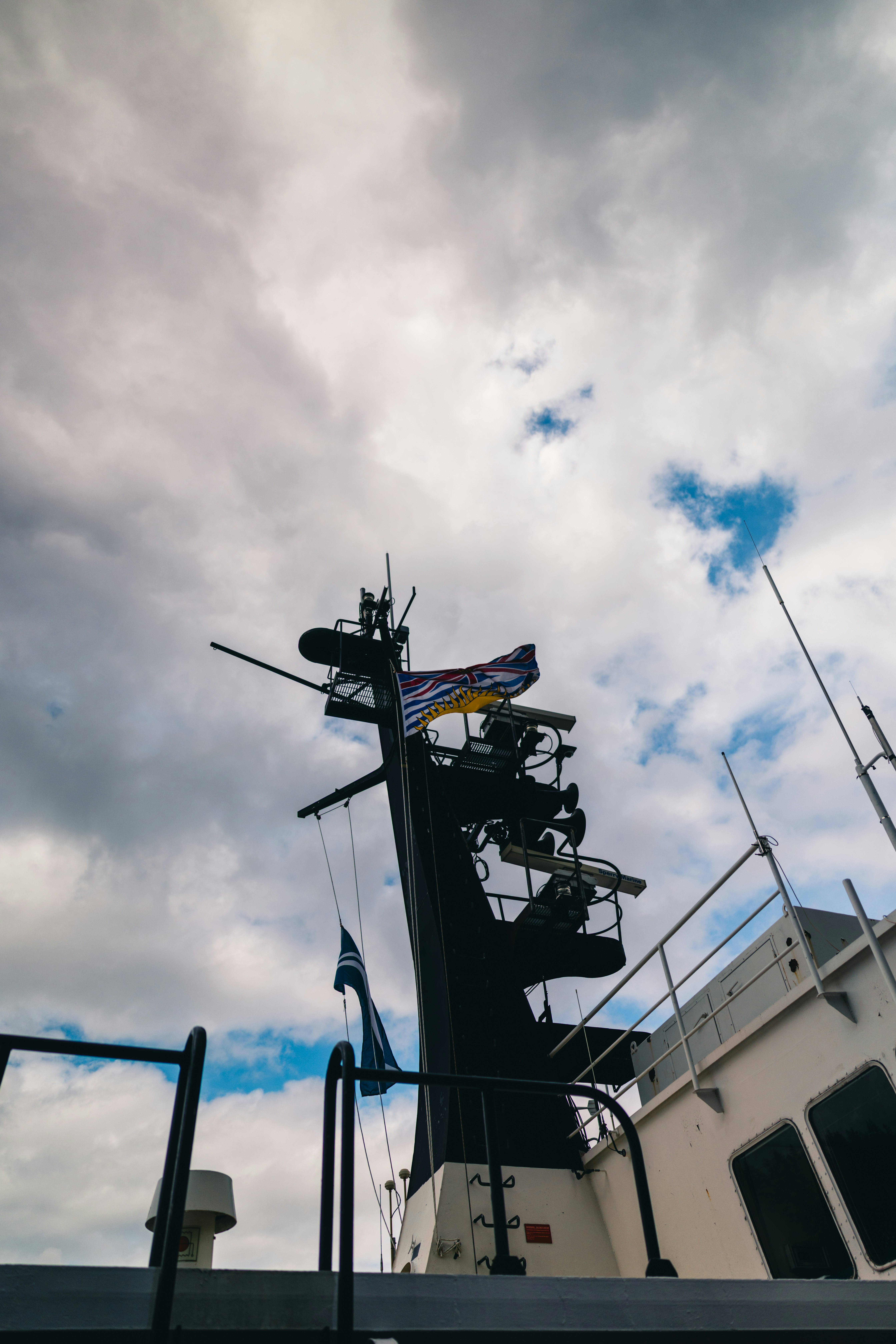 View of a ship's mast against a backdrop of dramatic clouds, showcasing nautical instruments and flags. The composition emphasizes the vessel's structure and the vastness of the sky.