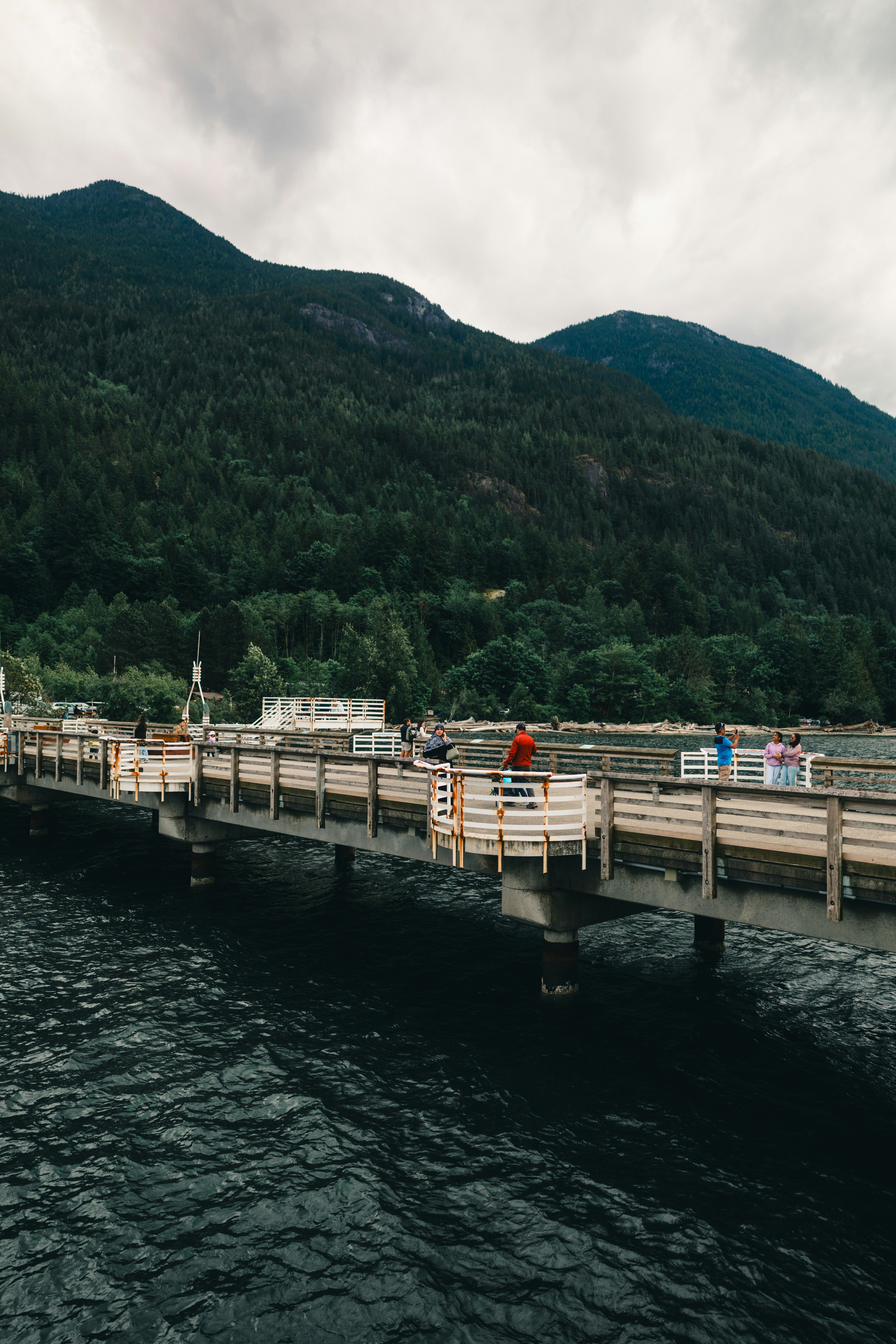 Wooden pier extending into calm waters, surrounded by lush green mountains under a cloudy sky. People enjoy the scenic view.