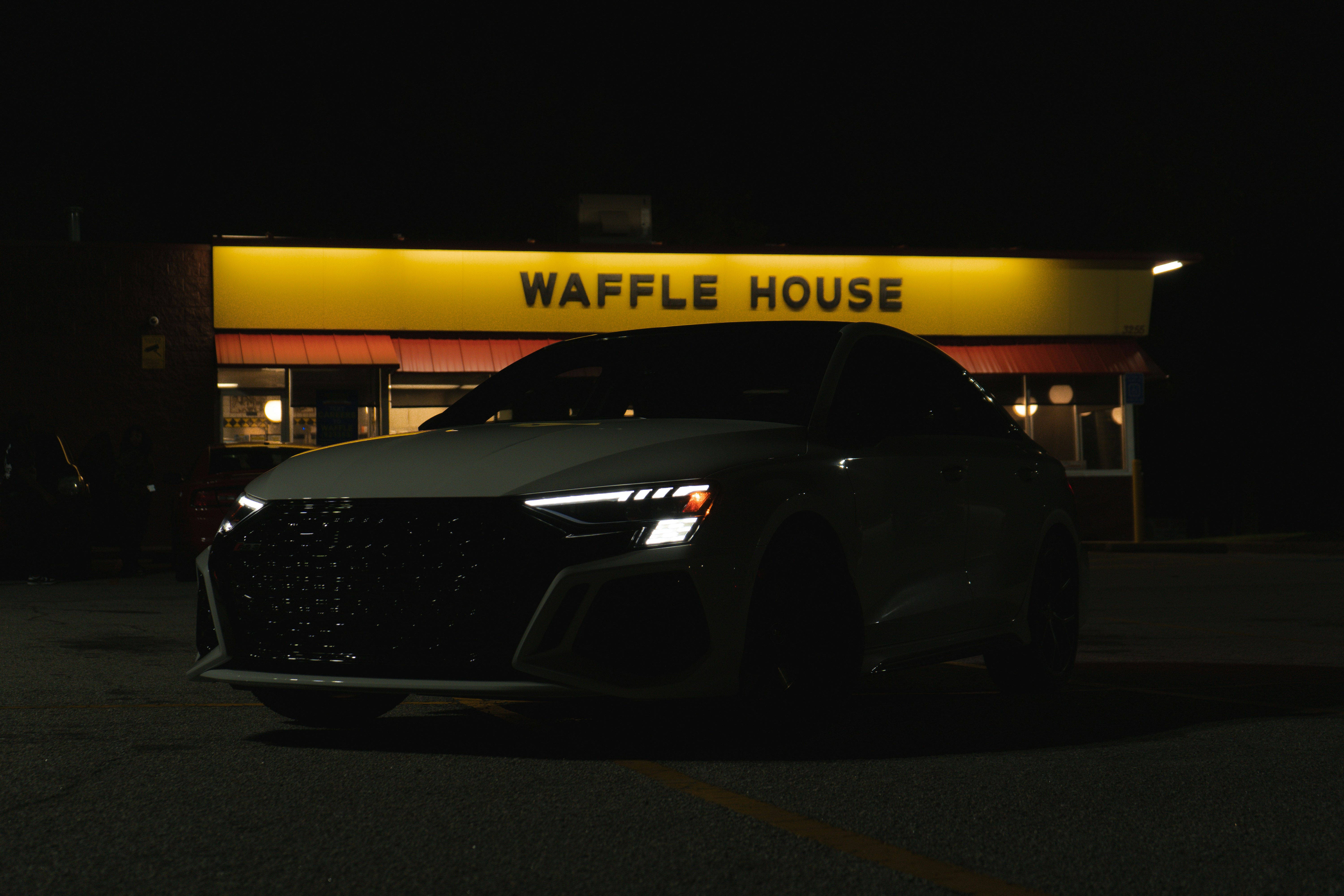 A sleek white car parked in front of a brightly lit Waffle House at night, highlighting the contrast between modern design and classic diner signage.
