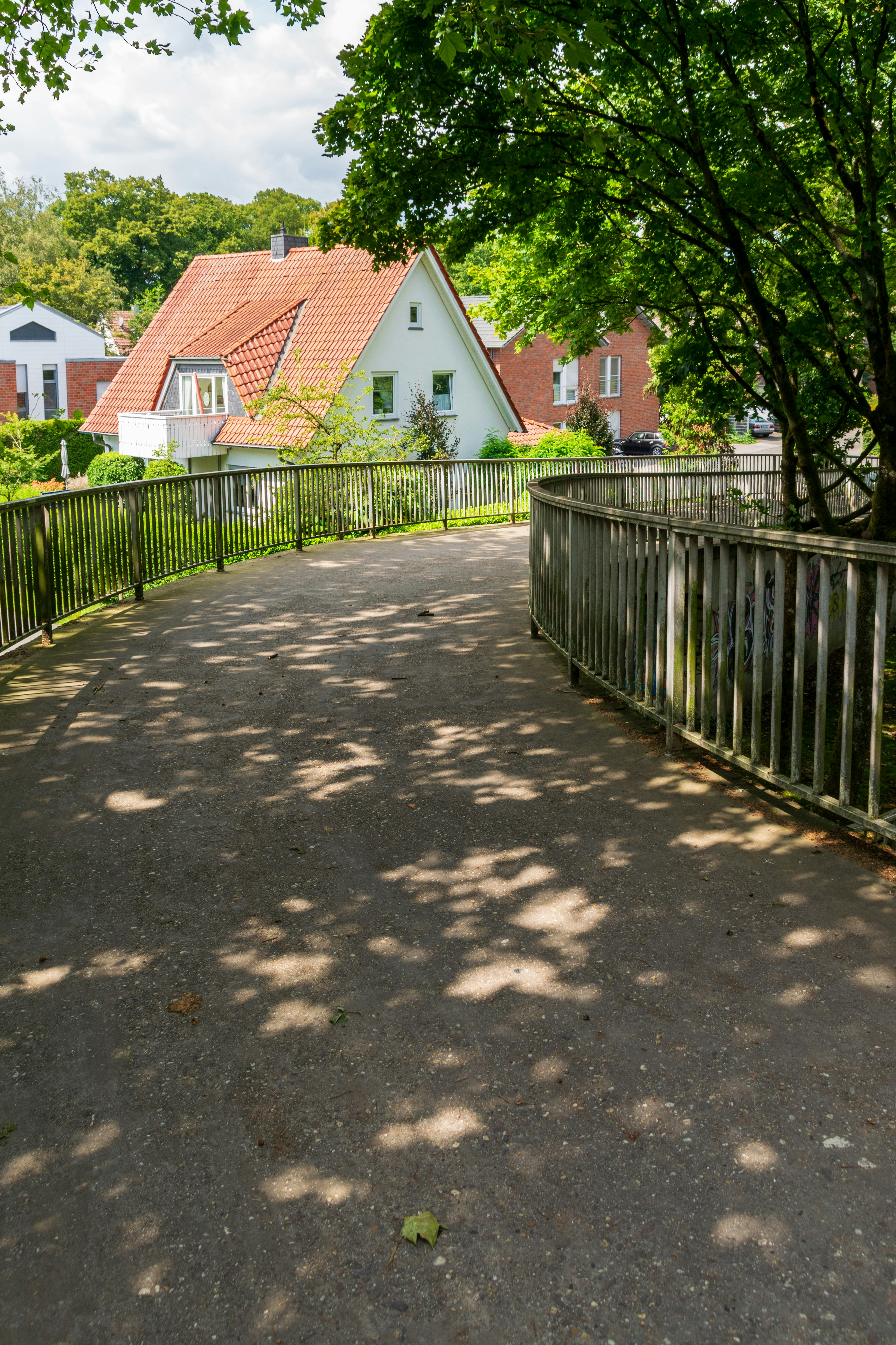 A winding pathway surrounded by lush greenery and shadows, leading towards a quaint house with a red-tiled roof.