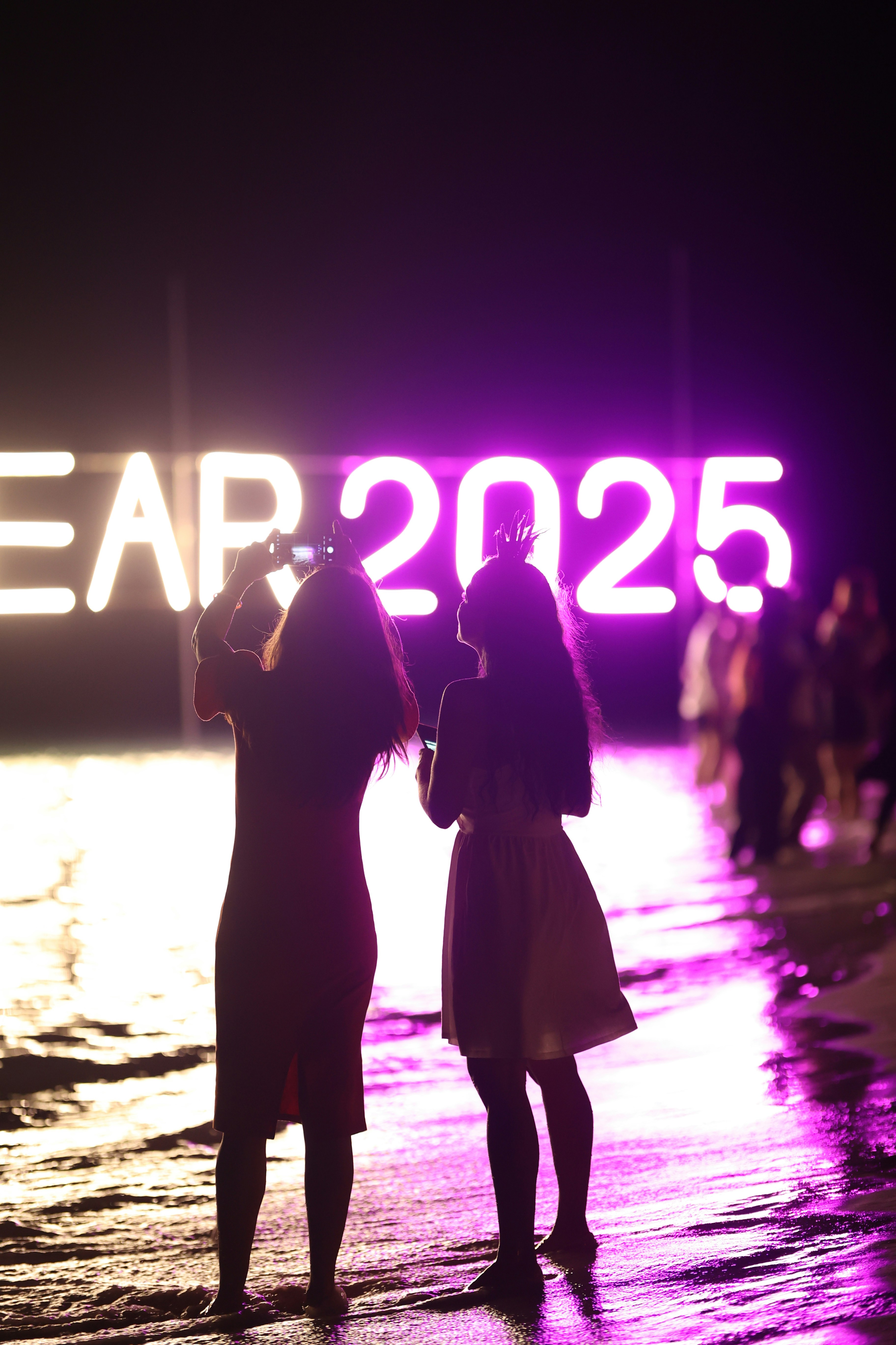 Silhouettes of two women capturing a moment at the beach with a vibrant 'YEAR 2025' sign illuminated in the background.