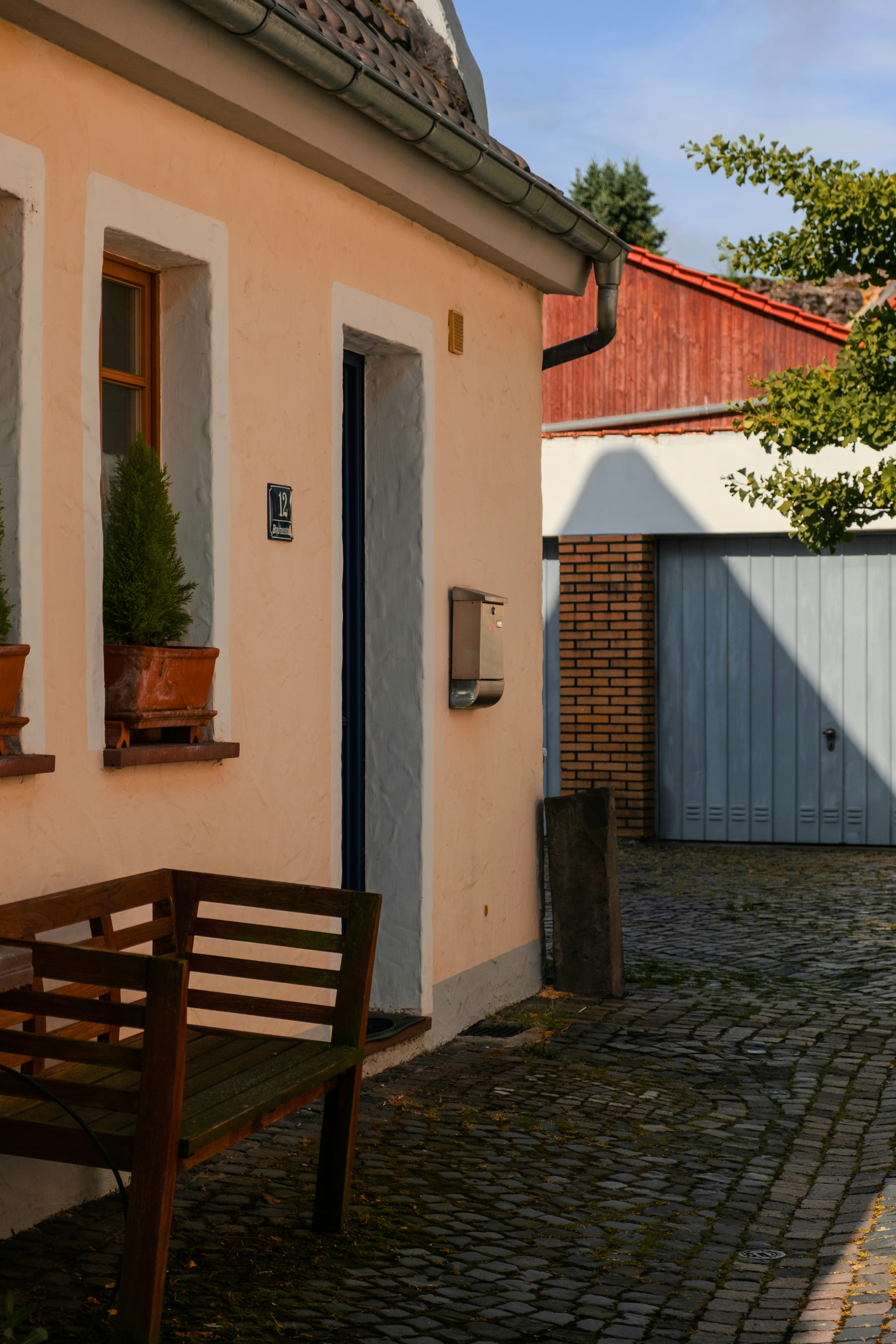Charming residential alley featuring a pastel-colored house with potted plants and a wooden bench, inviting relaxation. A glimpse of a garage and greenery adds to the serene atmosphere.