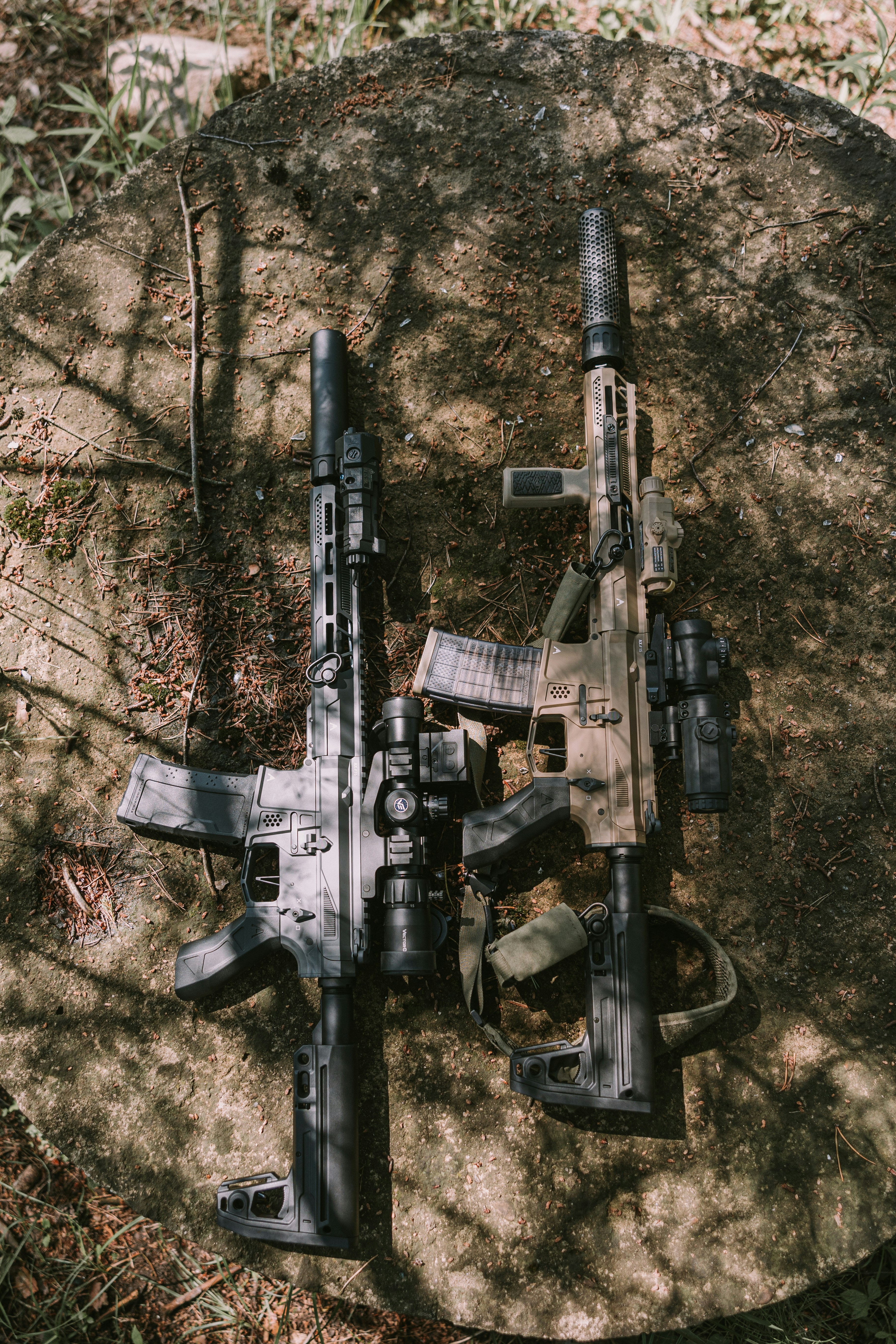 Two tactical rifles positioned on a stone surface, showcasing distinct designs and accessories under dappled sunlight.