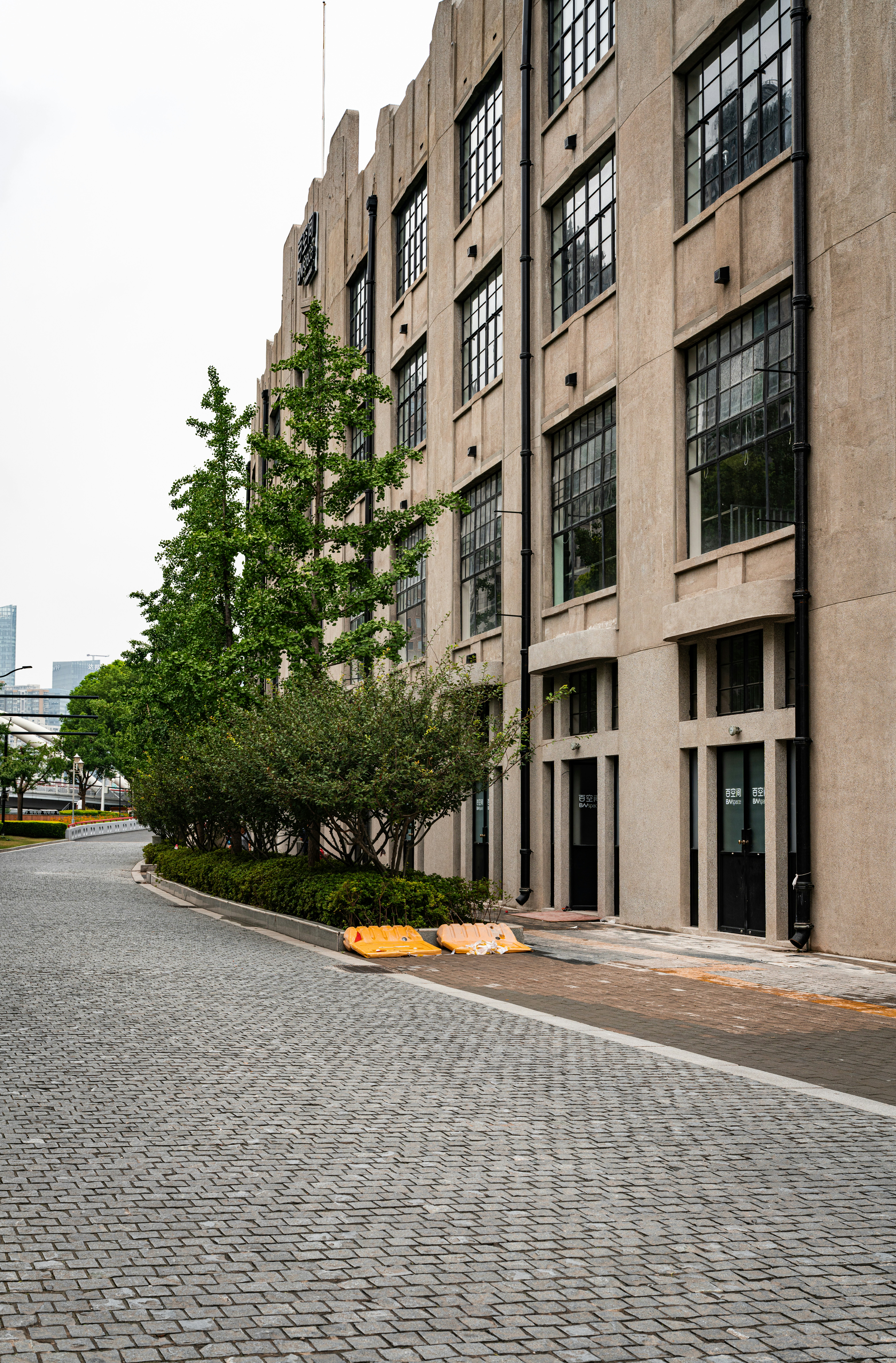 A modern industrial building framed by lush greenery along a cobblestone path, showcasing the blend of urban architecture and nature's resilience.