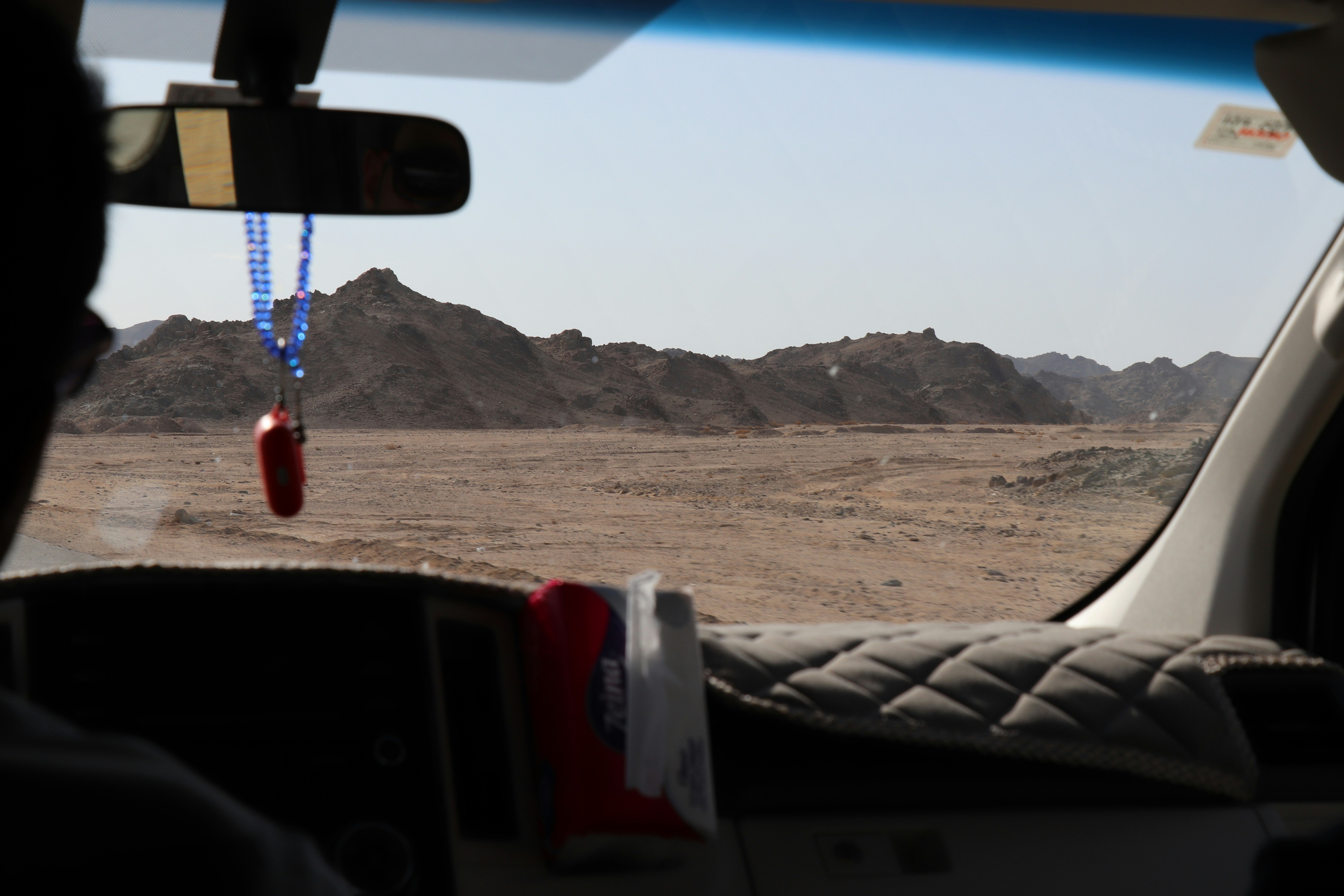 A raw and immersive shot of an Egyptian desert landscape seen from inside a car, capturing the spirit of adventure and road trips through barren, sun-scorched terrain. The dusty dashboard, the hanging charm, and the rugged mountains in the background all convey a sense of motion, solitude, and timelessness. Perfect for travel, exploration, and off-the-beaten-path themes. | Desert landscape viewed through a car windshield.