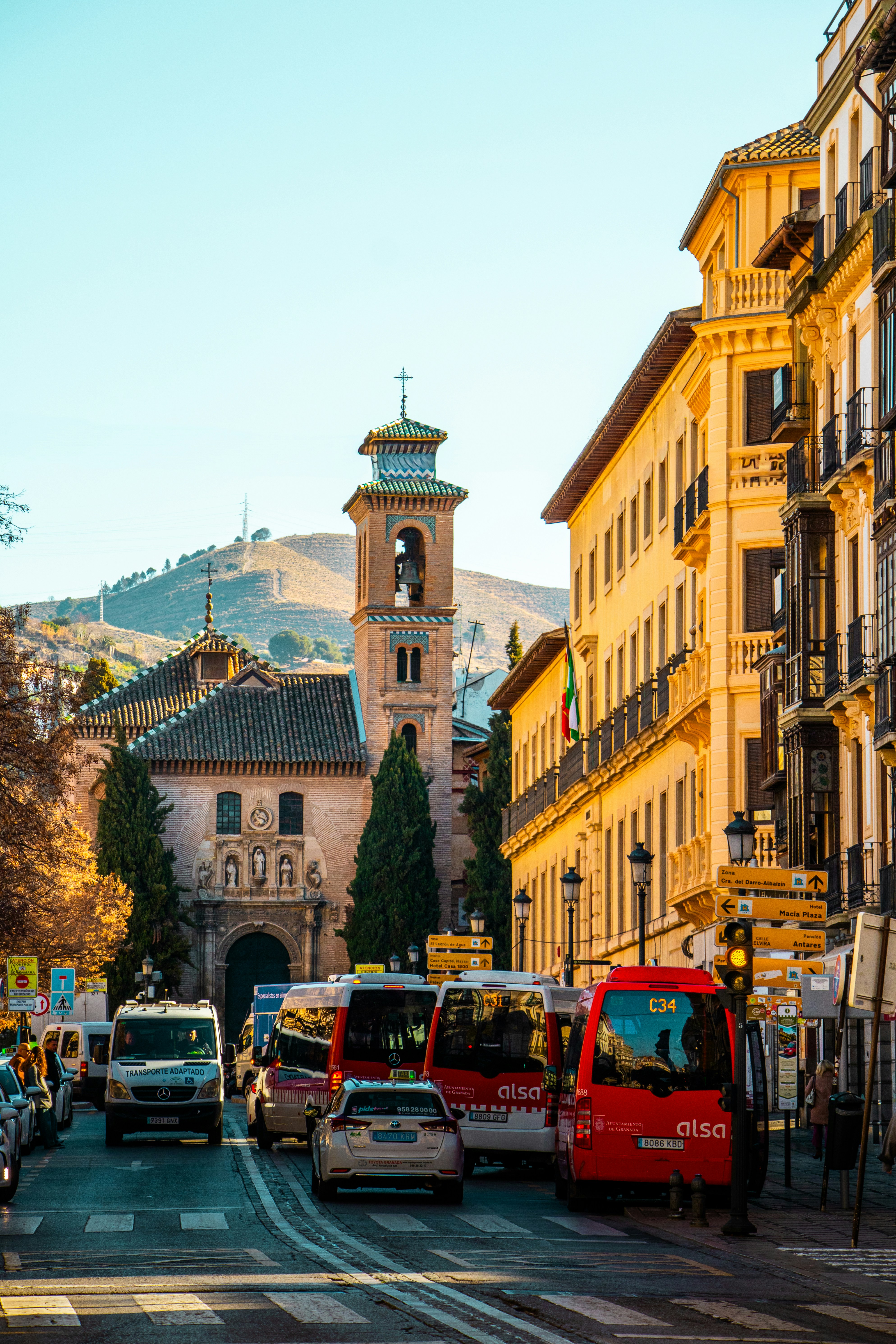 Historic buildings frame a bustling street scene in Granada, featuring the iconic bell tower and vibrant city buses. The backdrop showcases the scenic hills.