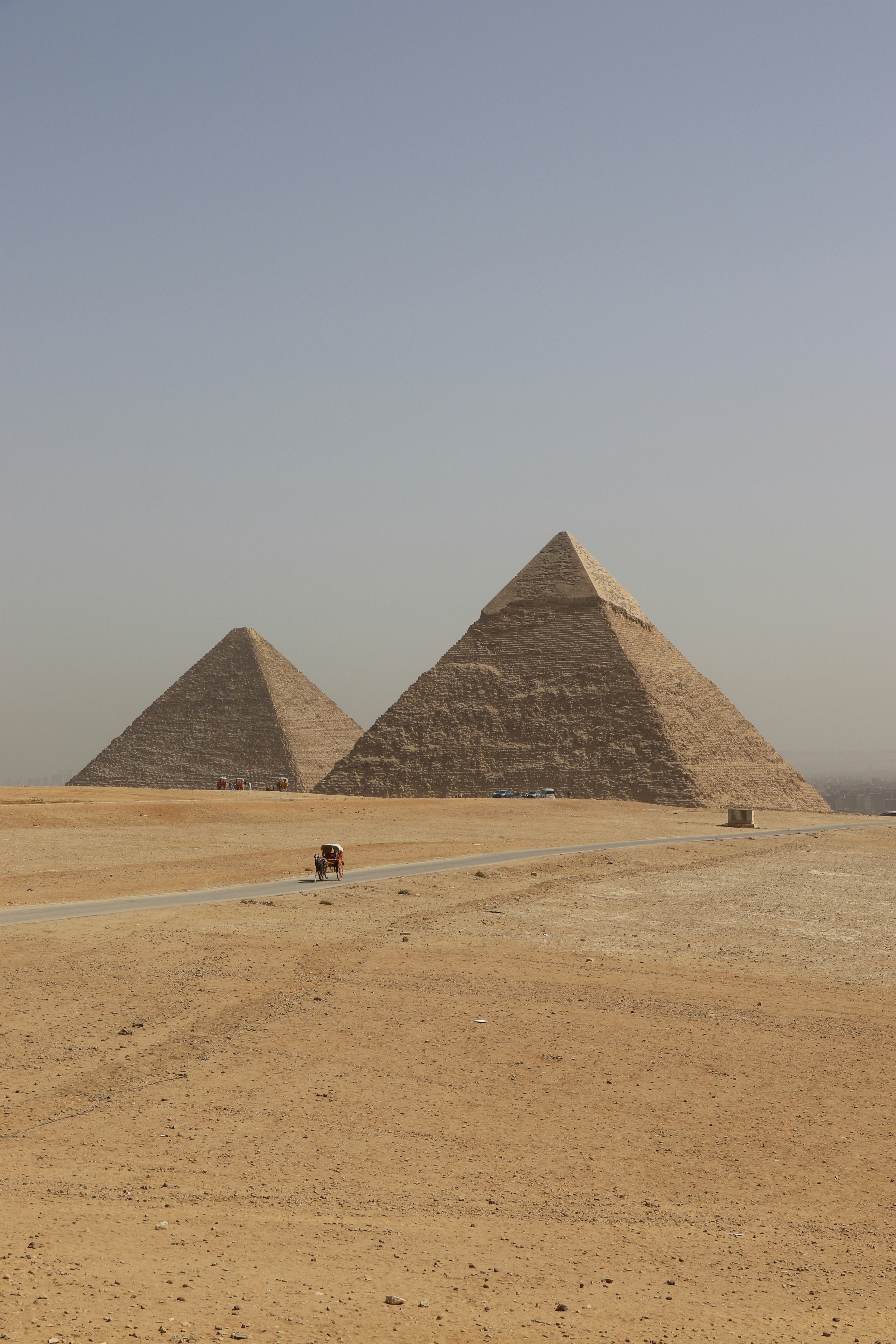 A timeless desert scene in Giza, Egypt, featuring two of the iconic pyramids standing tall under a pale blue sky. In the foreground, a traditional horse-drawn carriage makes its way through the arid landscape, offering a glimpse into the intersection of ancient grandeur and modern-day tourism. A powerful image of heritage, scale, and solitude in one of the world’s most legendary sites. | The pyramids of giza stand tall in egypt.