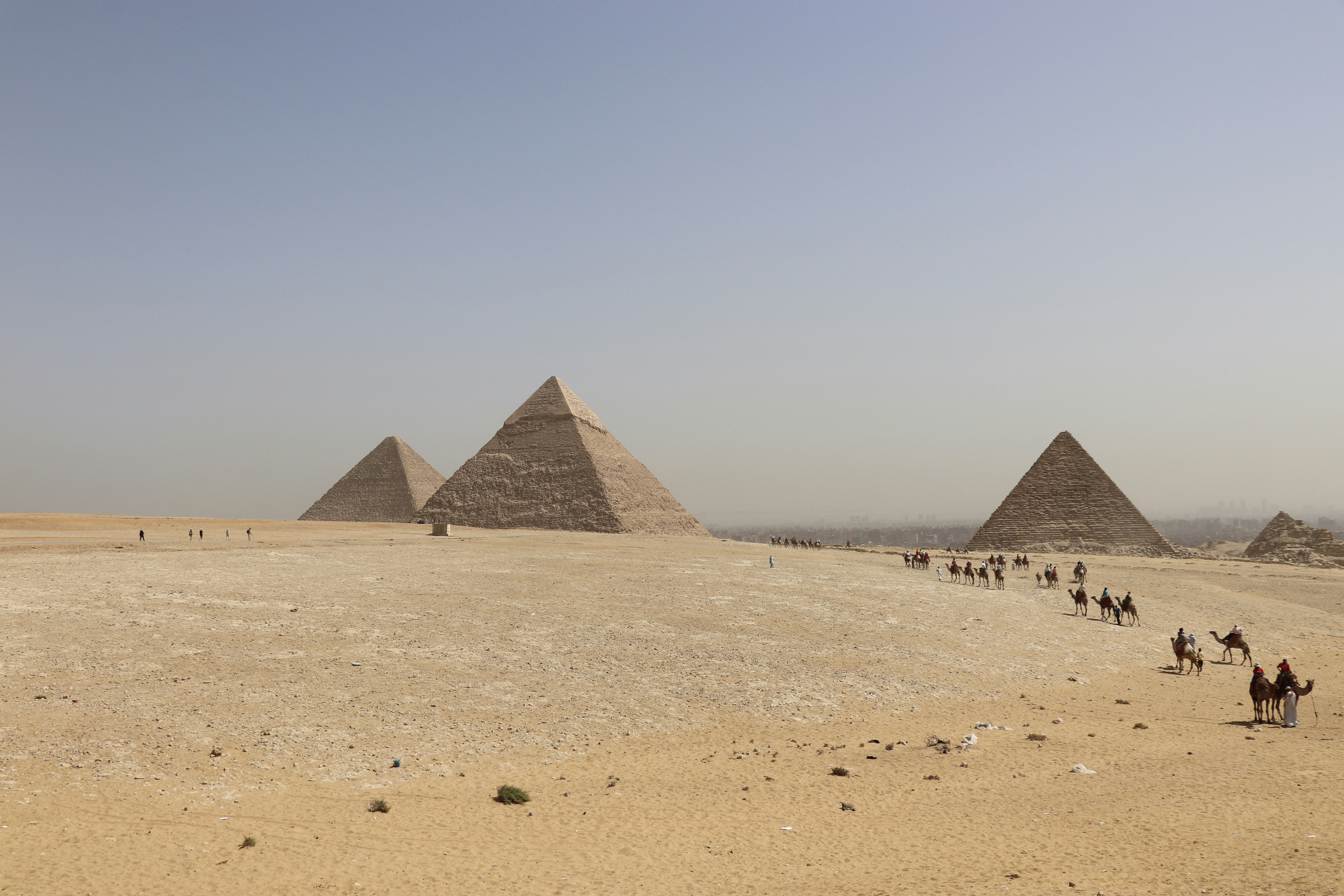A majestic view of the pyramids of Giza under a pale blue sky, with a long line of tourists riding camels across the golden desert. The soft haze in the background blends the ancient stone structures with the distant silhouette of Cairo. This shot captures the enduring charm of Egypt’s most iconic site, where history and culture meet timeless landscapes. | The great pyramids of giza stand tall in the desert.