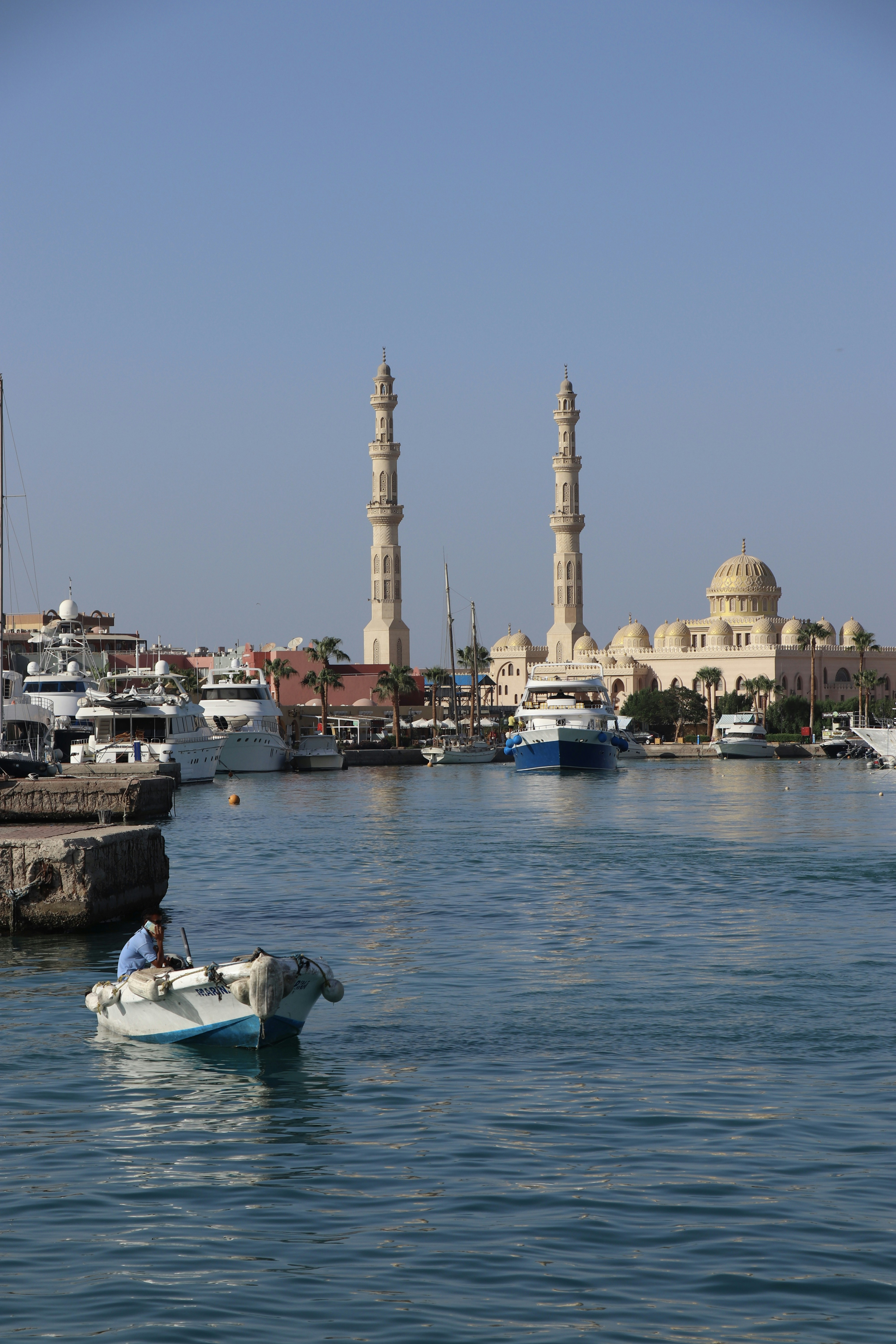 A peaceful view of the marina in Hurghada, Egypt, with the stunning Abdulmoneim Riadh Mosque rising in the background. The soft blue sky and calm waters reflect the harmony of daily life by the Red Sea. A lone fisherman in a small boat contrasts with the luxury yachts and domed architecture, capturing a moment where tradition and modernity meet on the Egyptian coast. | A boat floats in front of a scenic mosque.